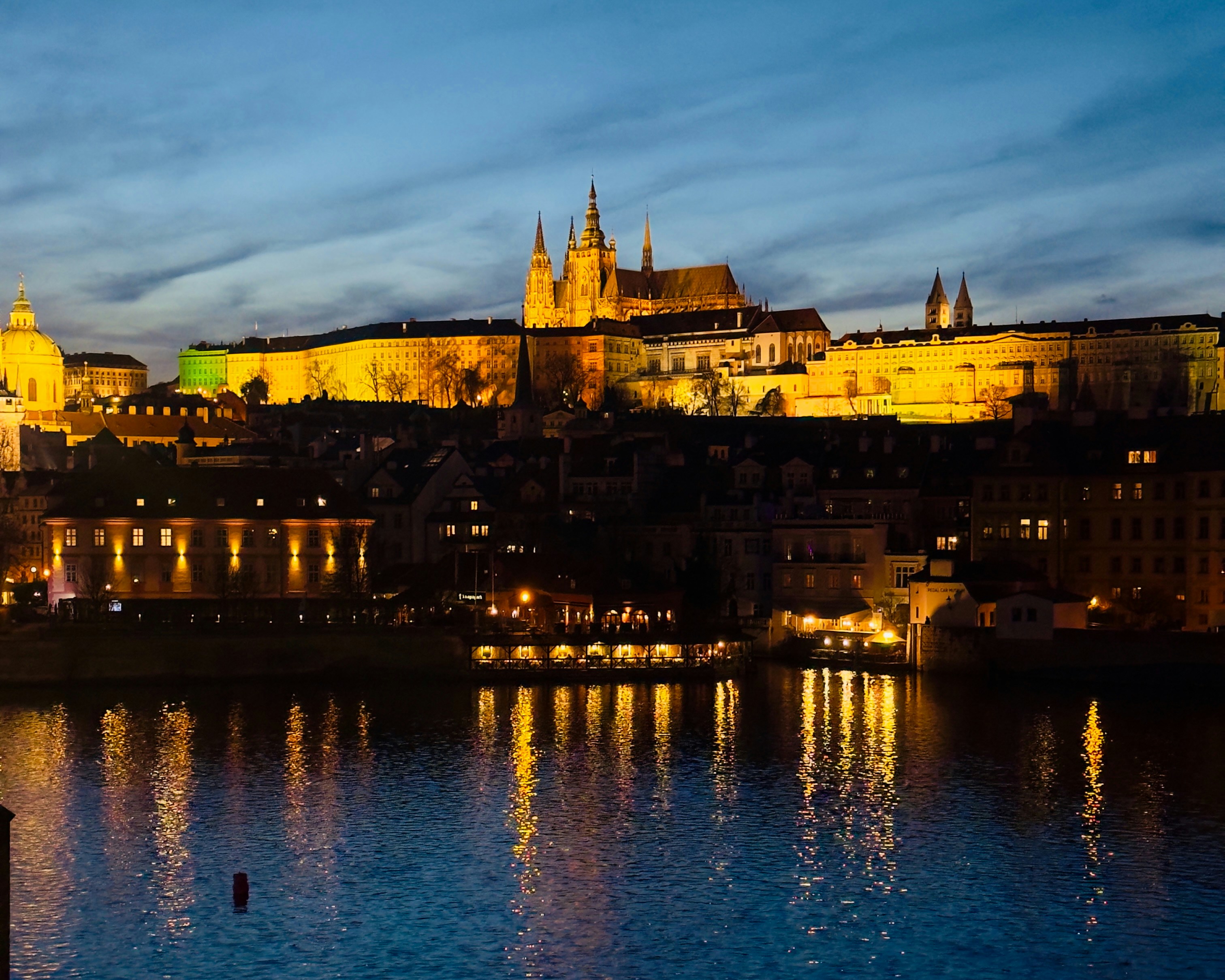 Prague Castle illuminated in warm tones, mirrored in the calm waters of the Vltava River under a twilight sky.
