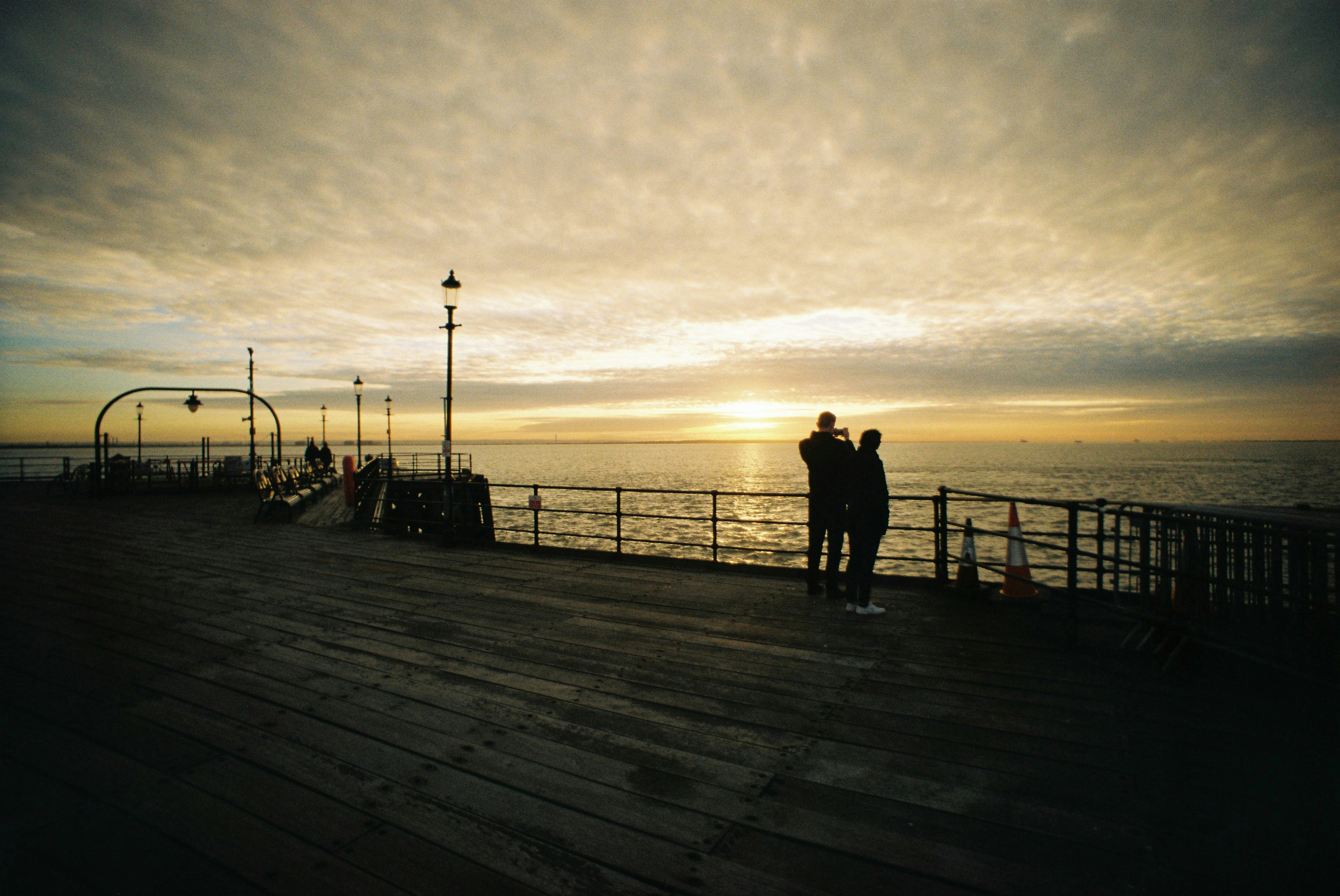Silhouetted couple stands on a pier with a vast ocean and golden sunset sky.