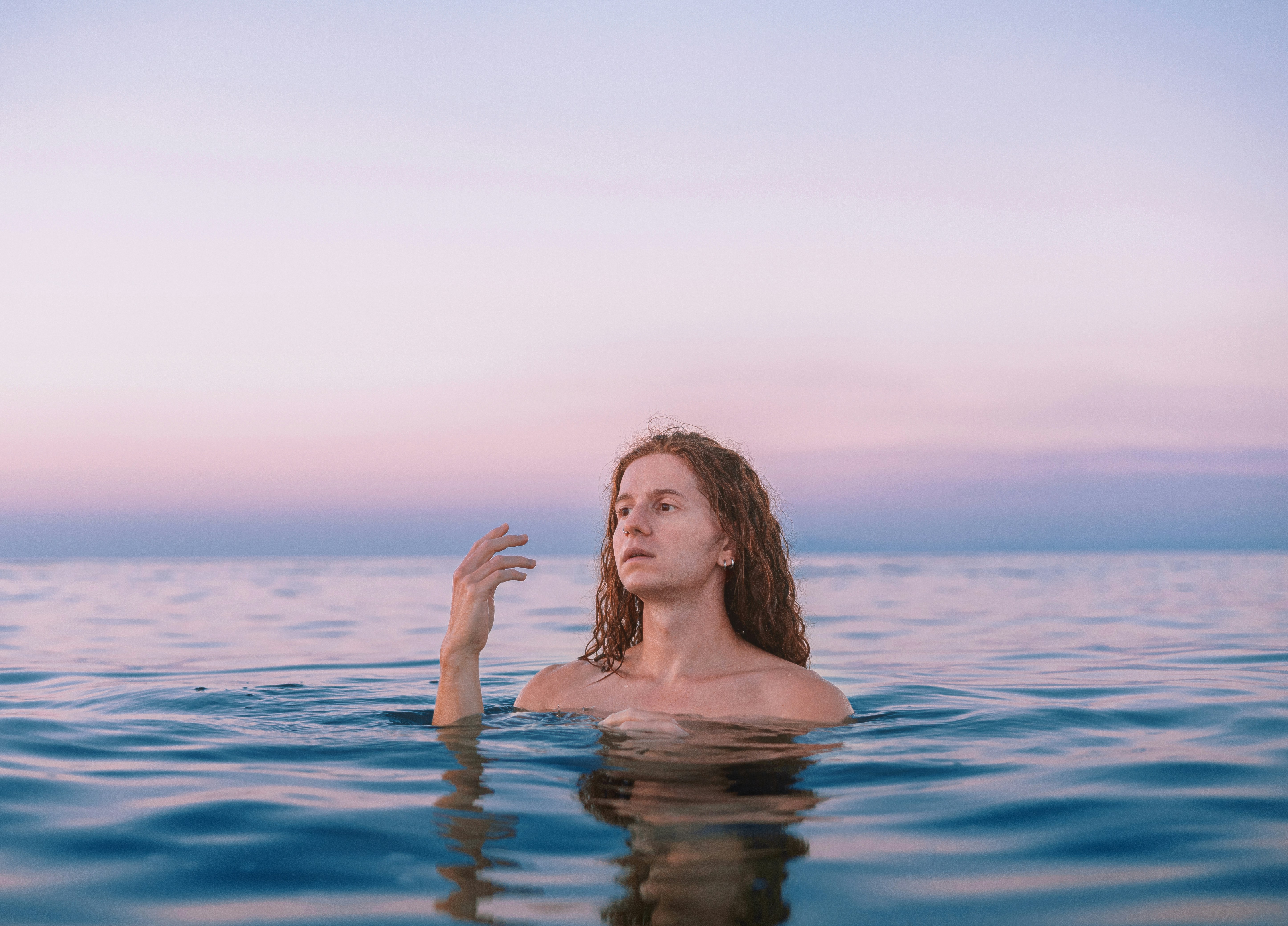 Person with long hair in calm ocean waters under a pastel sunset sky.