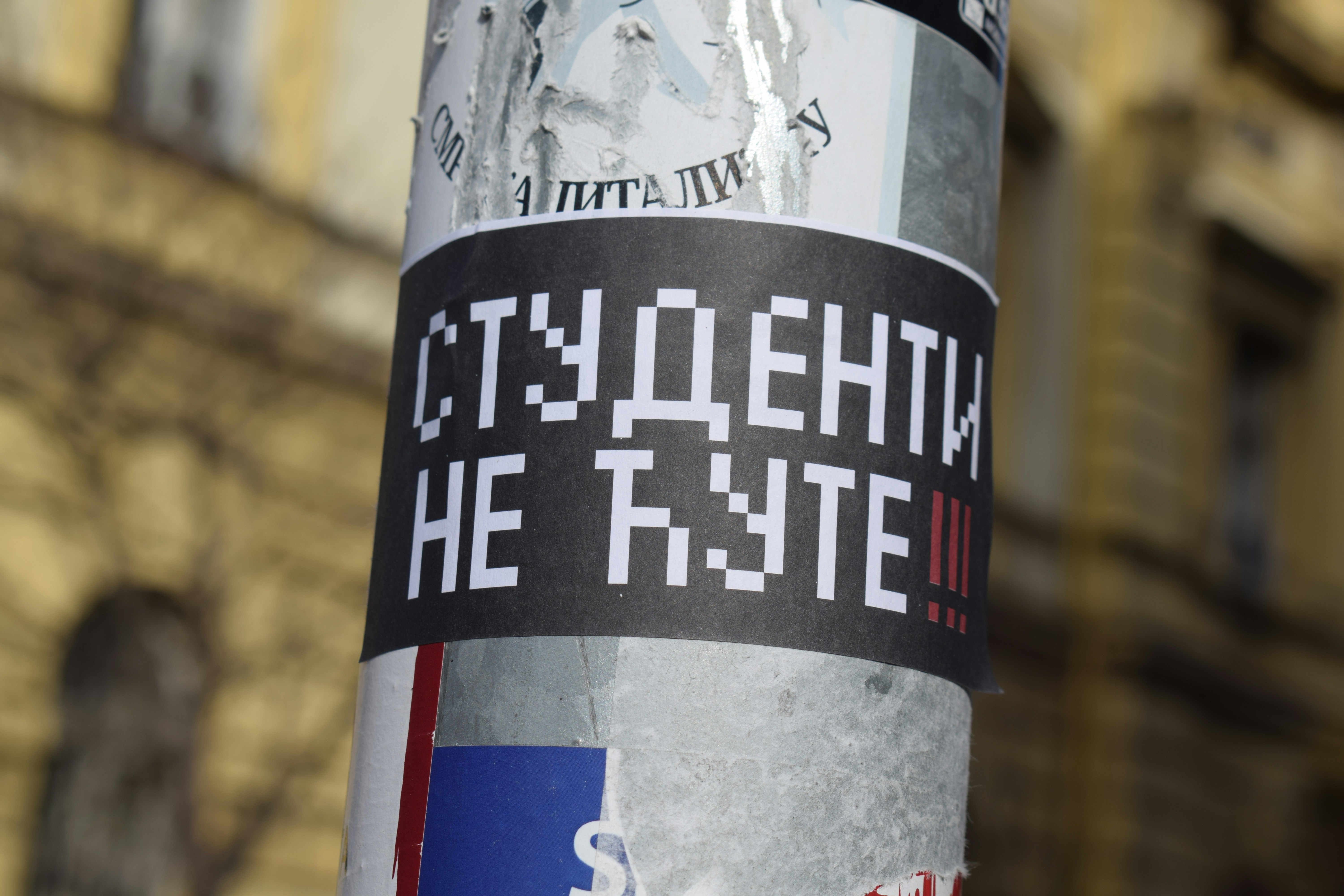 A striking black-and-white sticker on a pole, featuring bold text urging students not to be misled. The urban backdrop hints at a lively city environment.