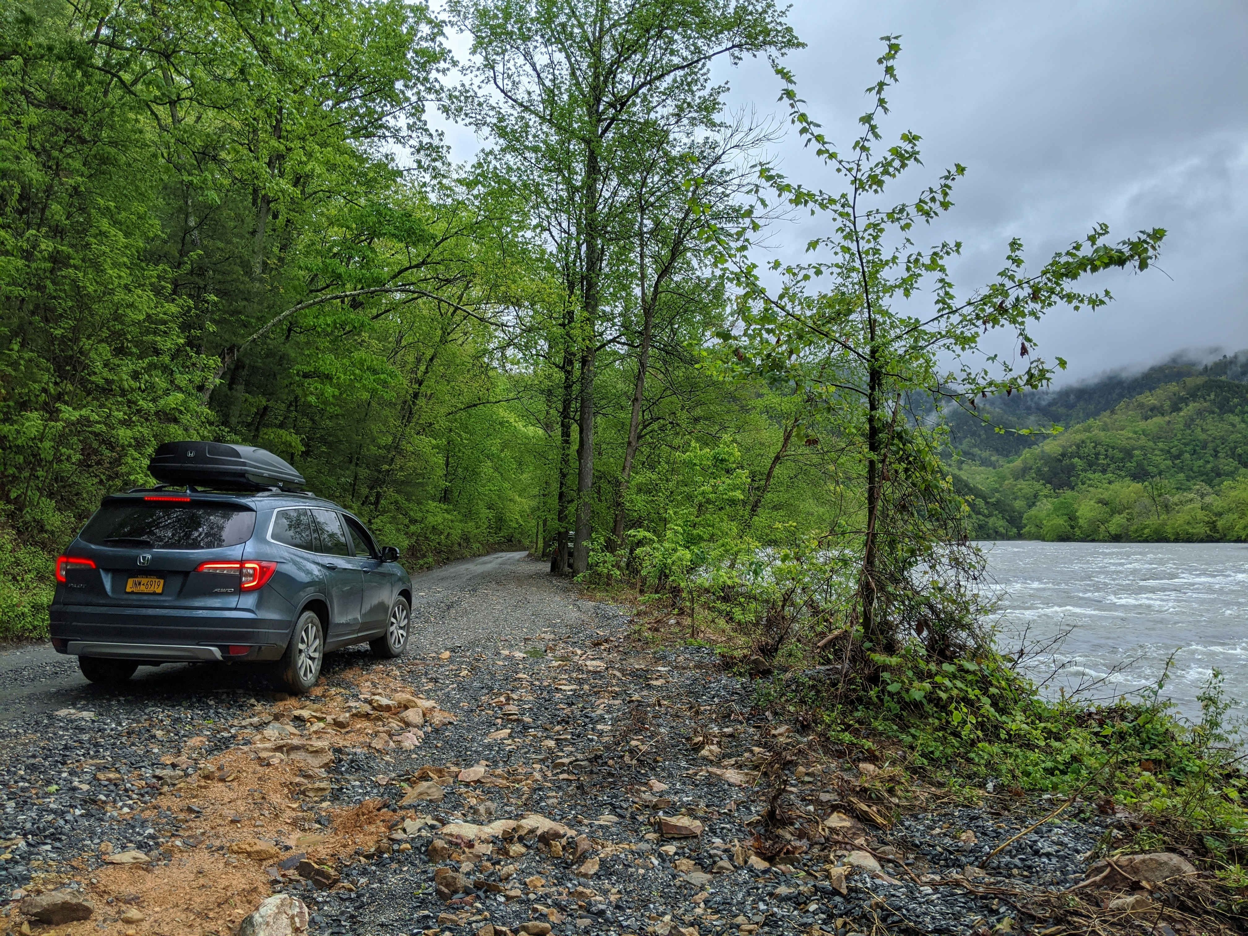 Blue SUV with a rooftop cargo box parked on a rocky riverside trail, lush green trees along the bank, and a fast-flowing river under a cloudy sky.