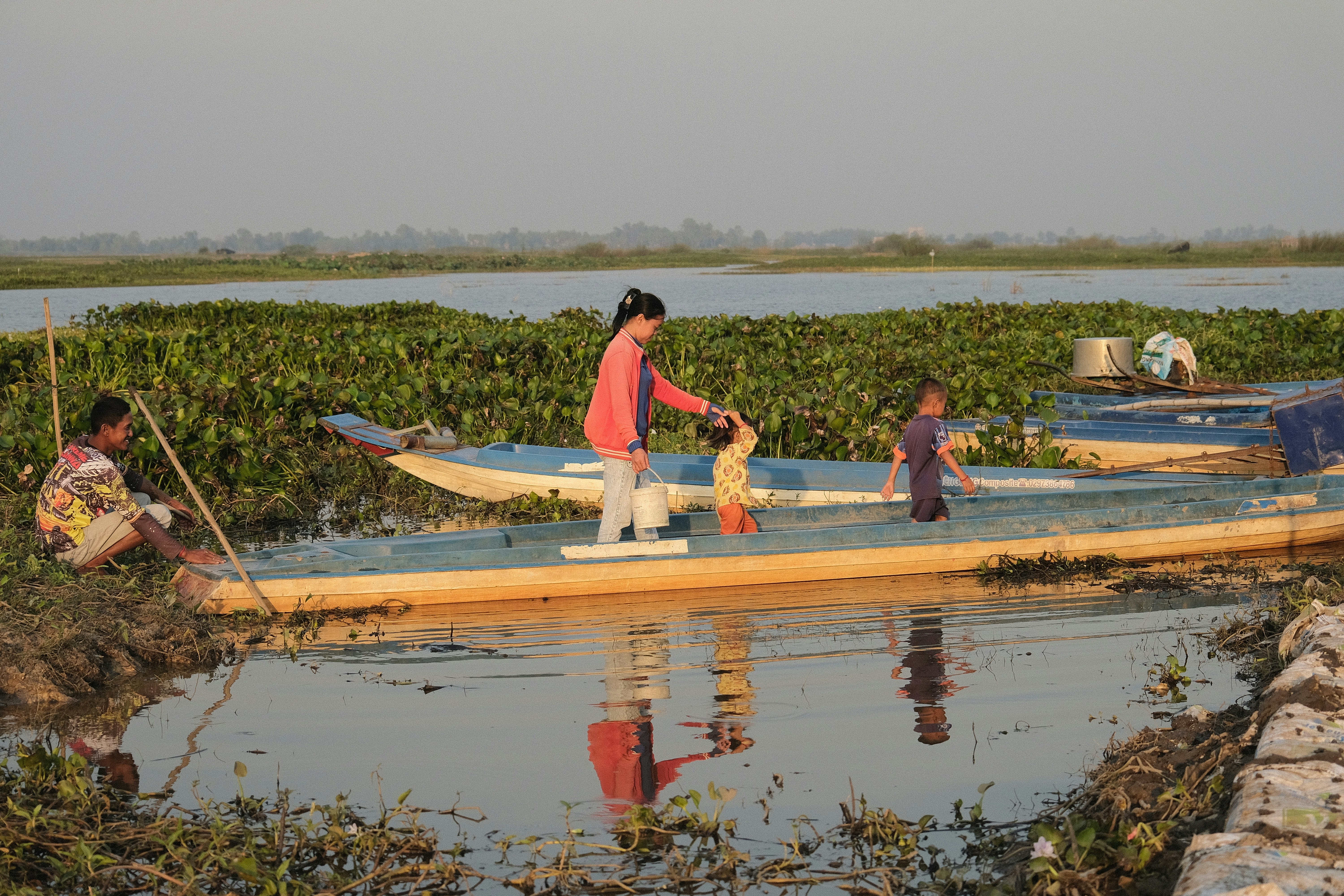 Family navigating a narrow wooden boat through lush water vegetation on a tranquil river.