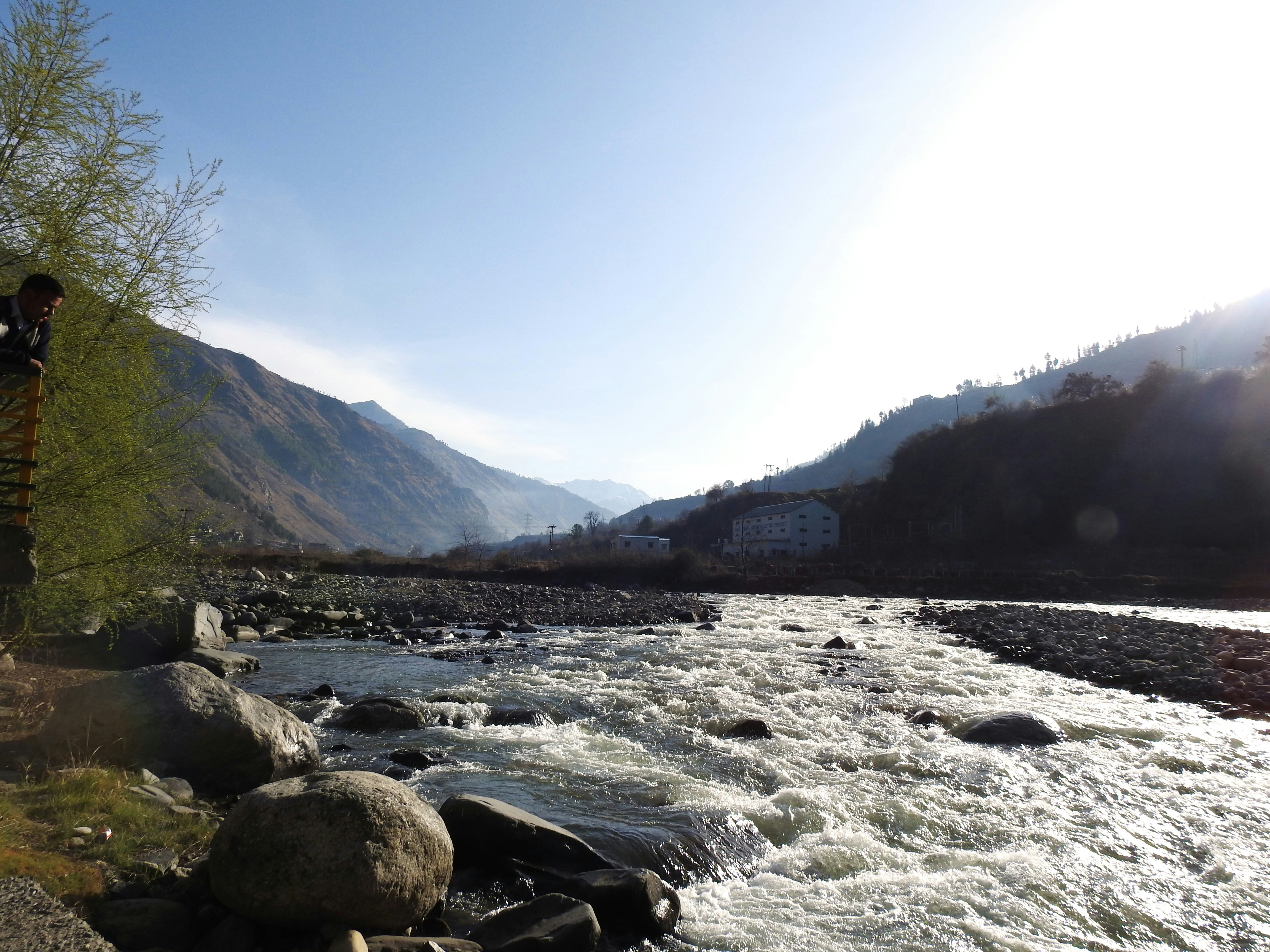 Man on a rock gazing at a river flowing through a mountainous landscape under a bright sky.