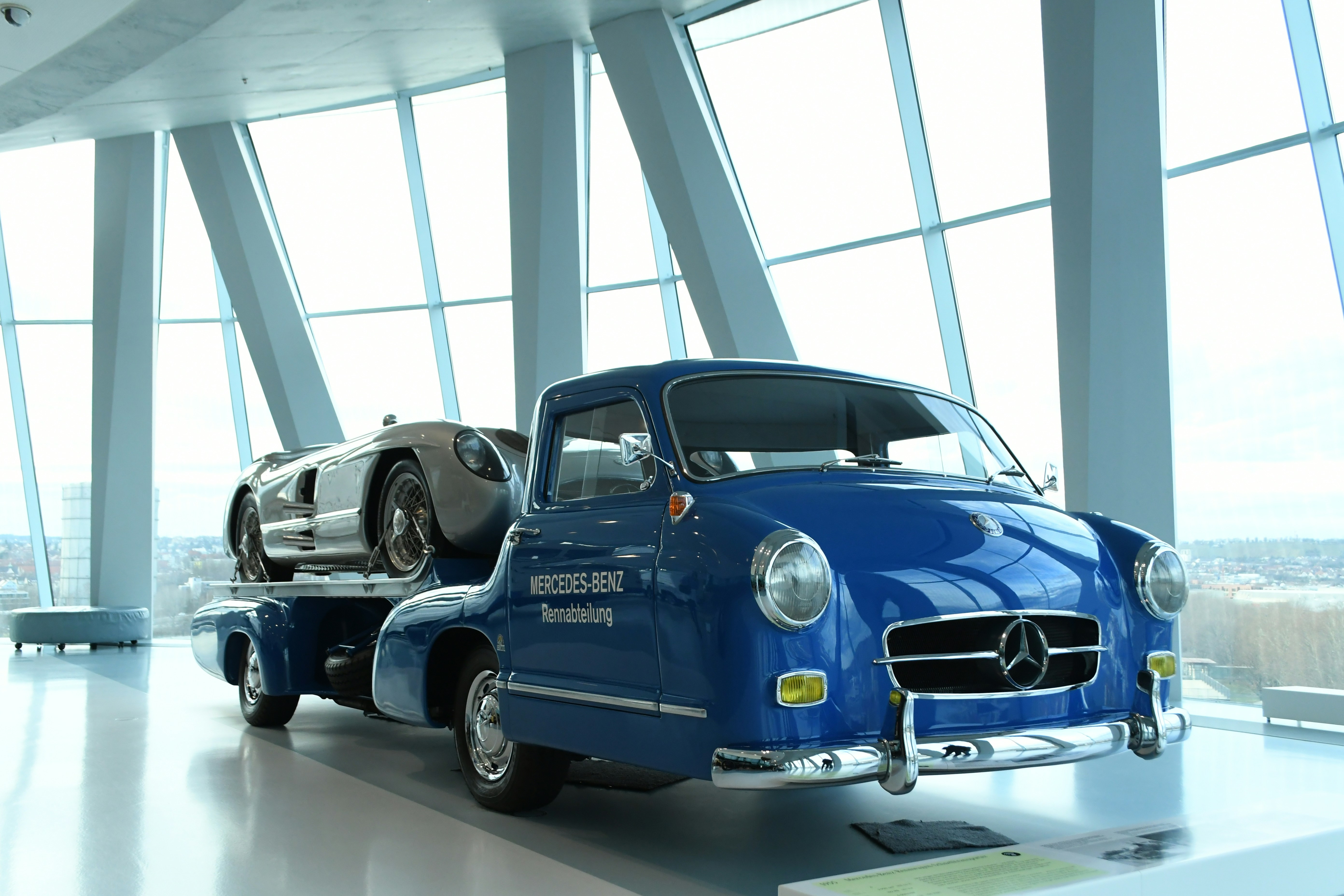 Vintage blue Mercedes-Benz transporter carrying a classic silver race car inside a modern glass-walled showroom.