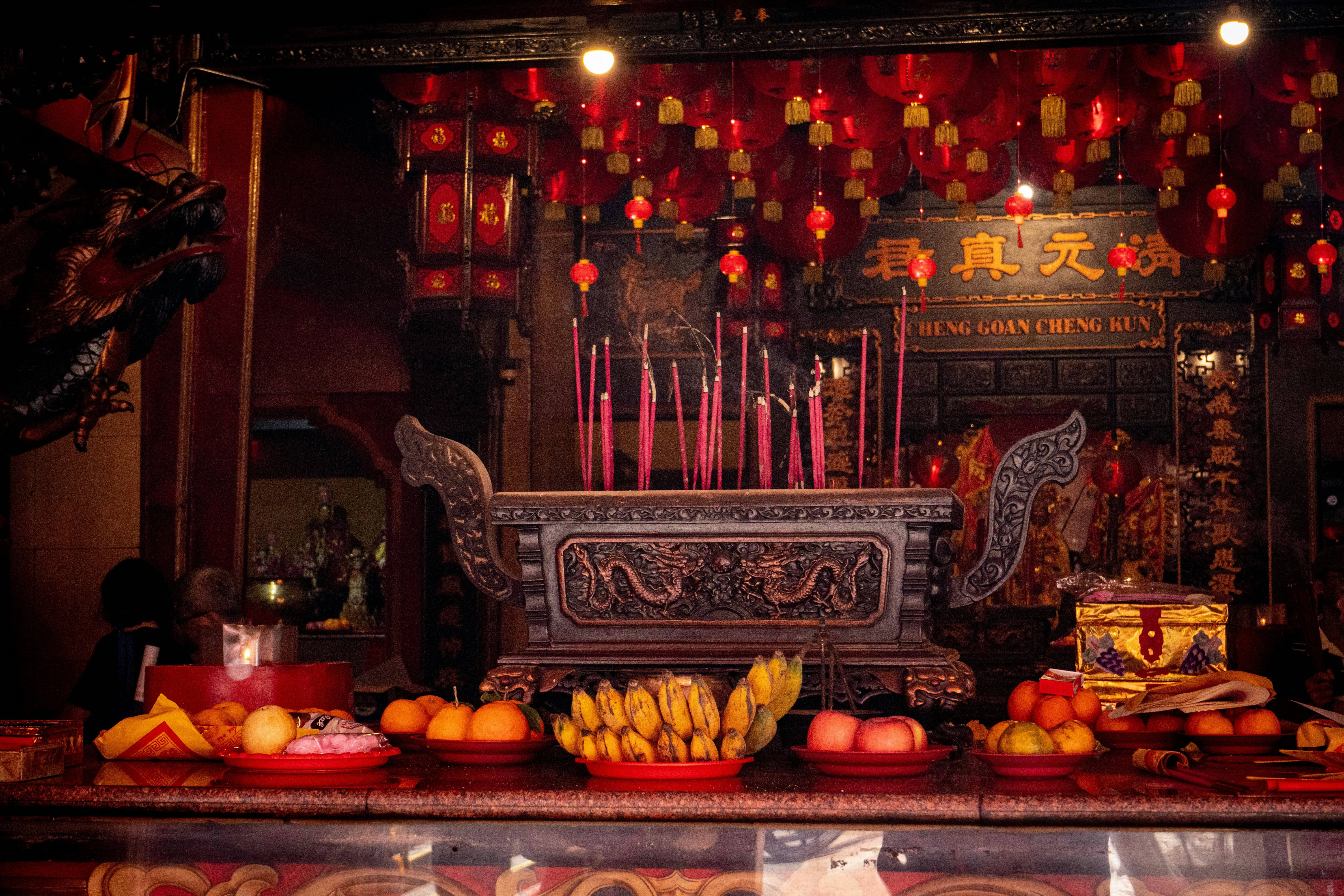 Incense sticks and vibrant fruits arranged on an ornate altar in a temple, surrounded by red lanterns and intricate decorations.