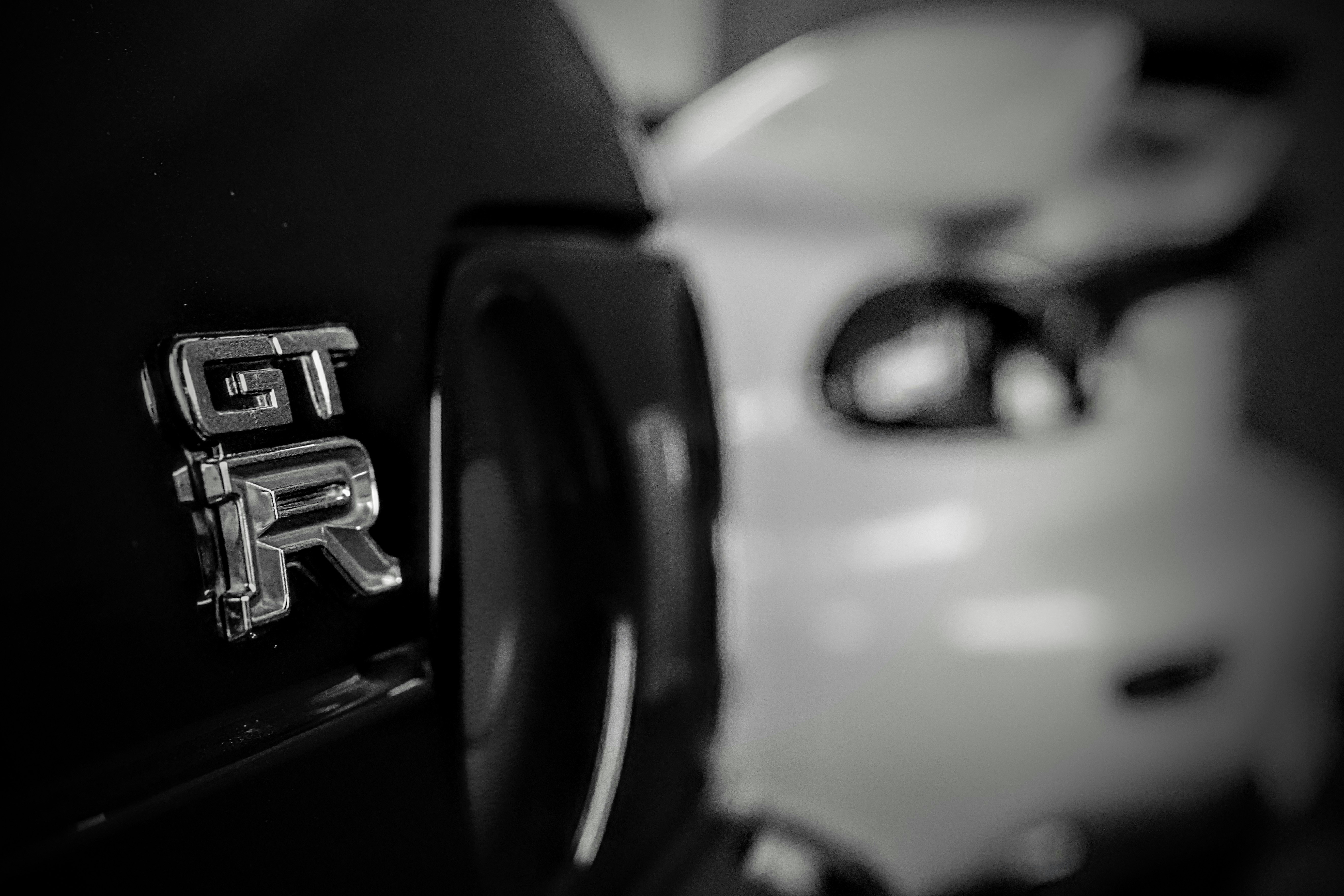 Macro close-up of the Toyota GR badge with a blurred white tail in monochrome