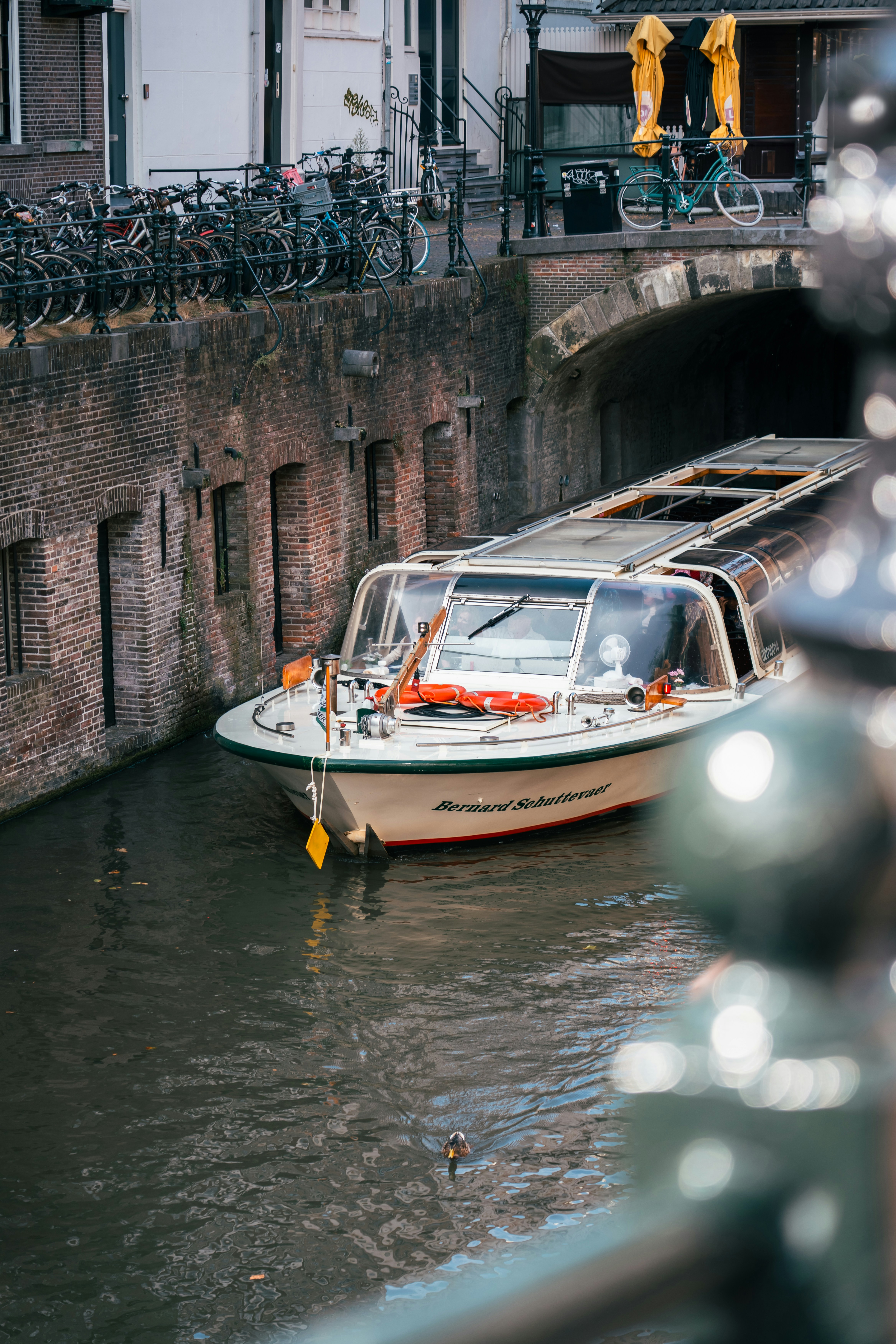Tour boat navigating a narrow canal with bicycles lined along the bridge above.