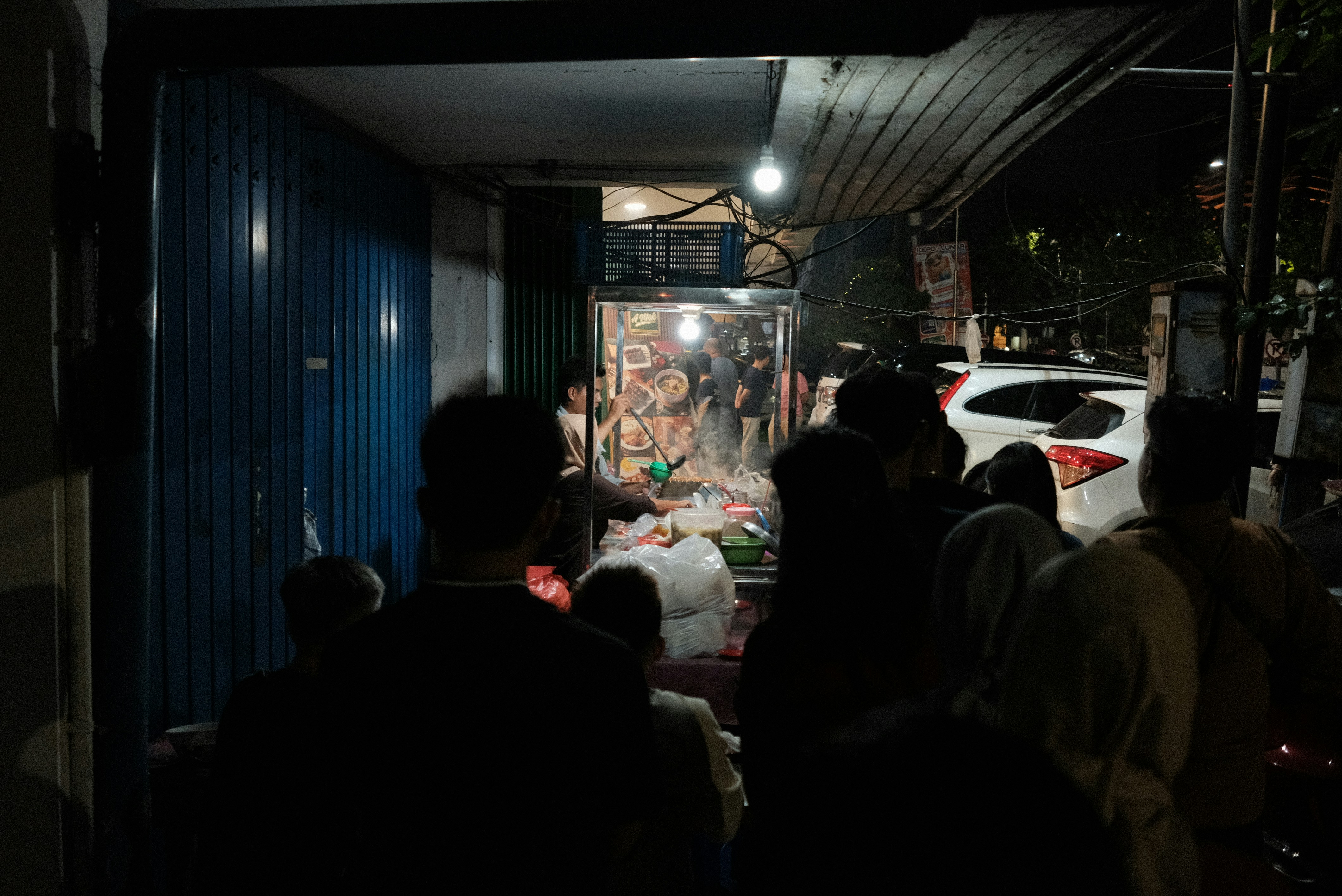 Street food stall bustling with activity at night, illuminated by an overhead bulb. The warm light highlights the vendor and eager customers in a dark alley, creating an intimate atmosphere.