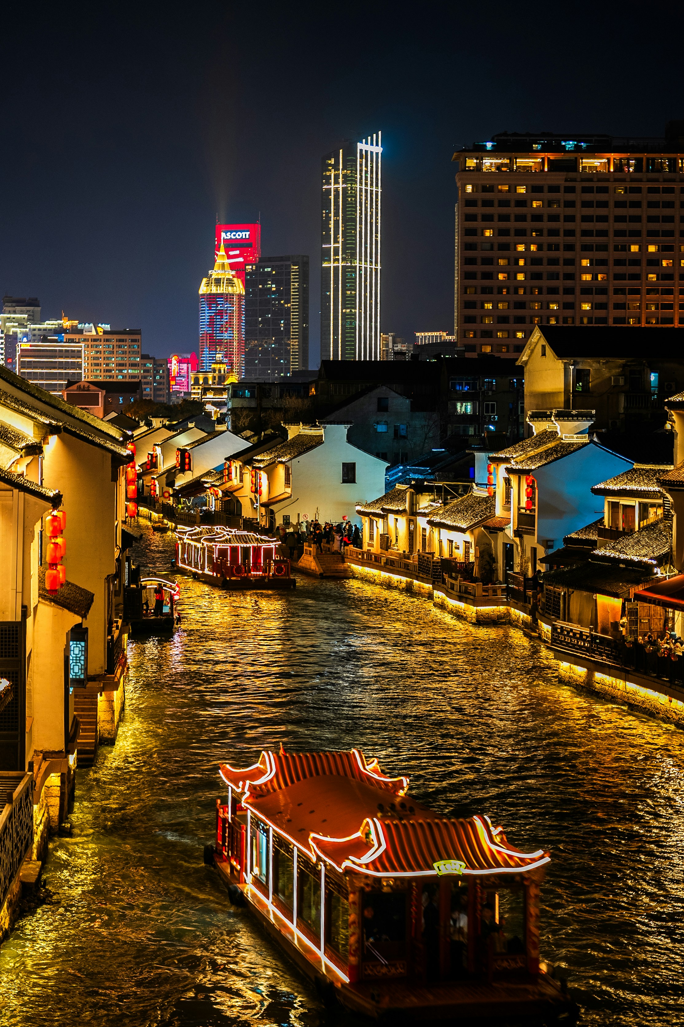 A vibrant scene of traditional boats gliding through a canal lined with illuminated buildings, showcasing the contrast between historic architecture and modern skyscrapers at night.