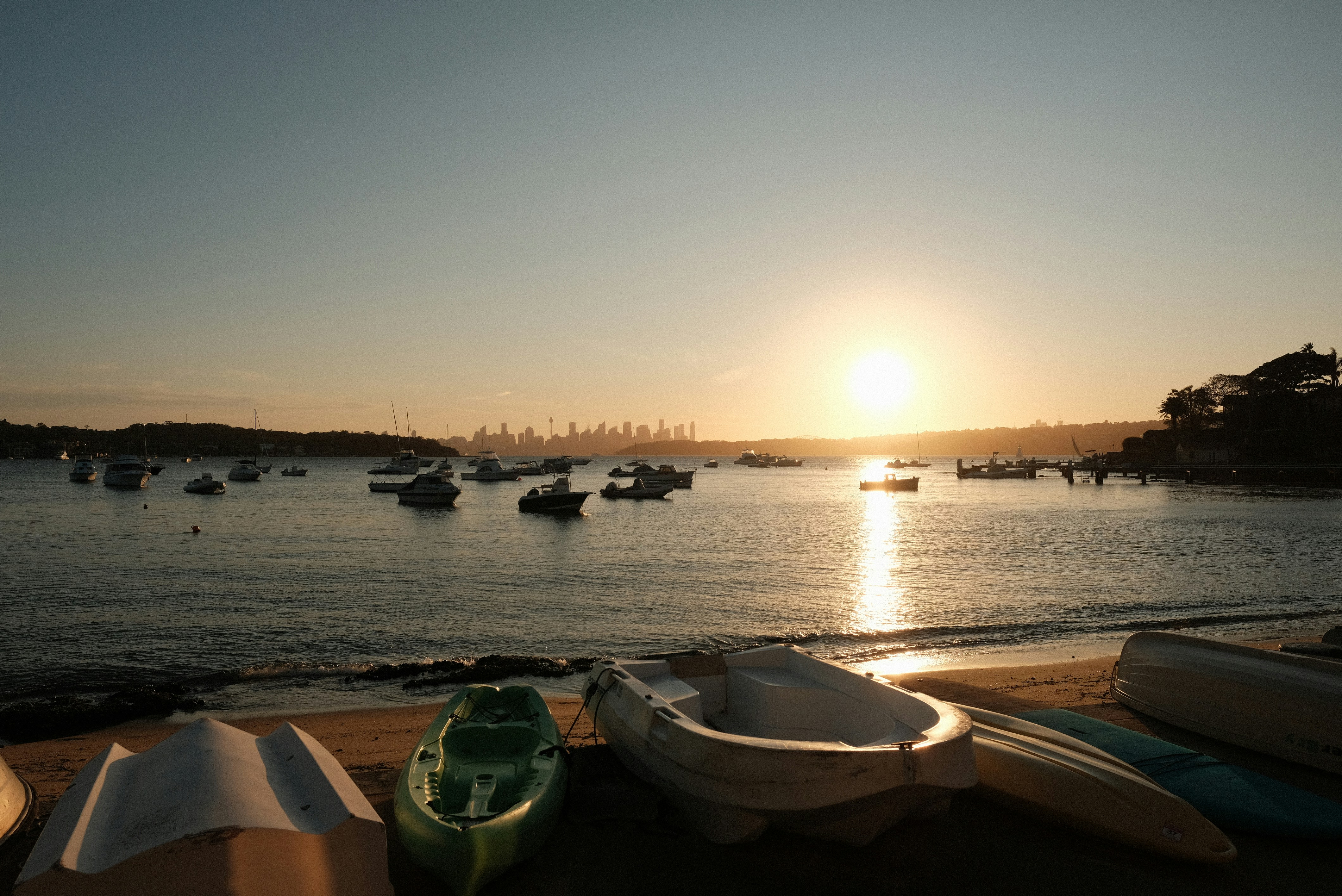 Sunset casts a warm glow over a tranquil harbor with boats anchored in calm waters and colorful vessels on the sandy shore.
