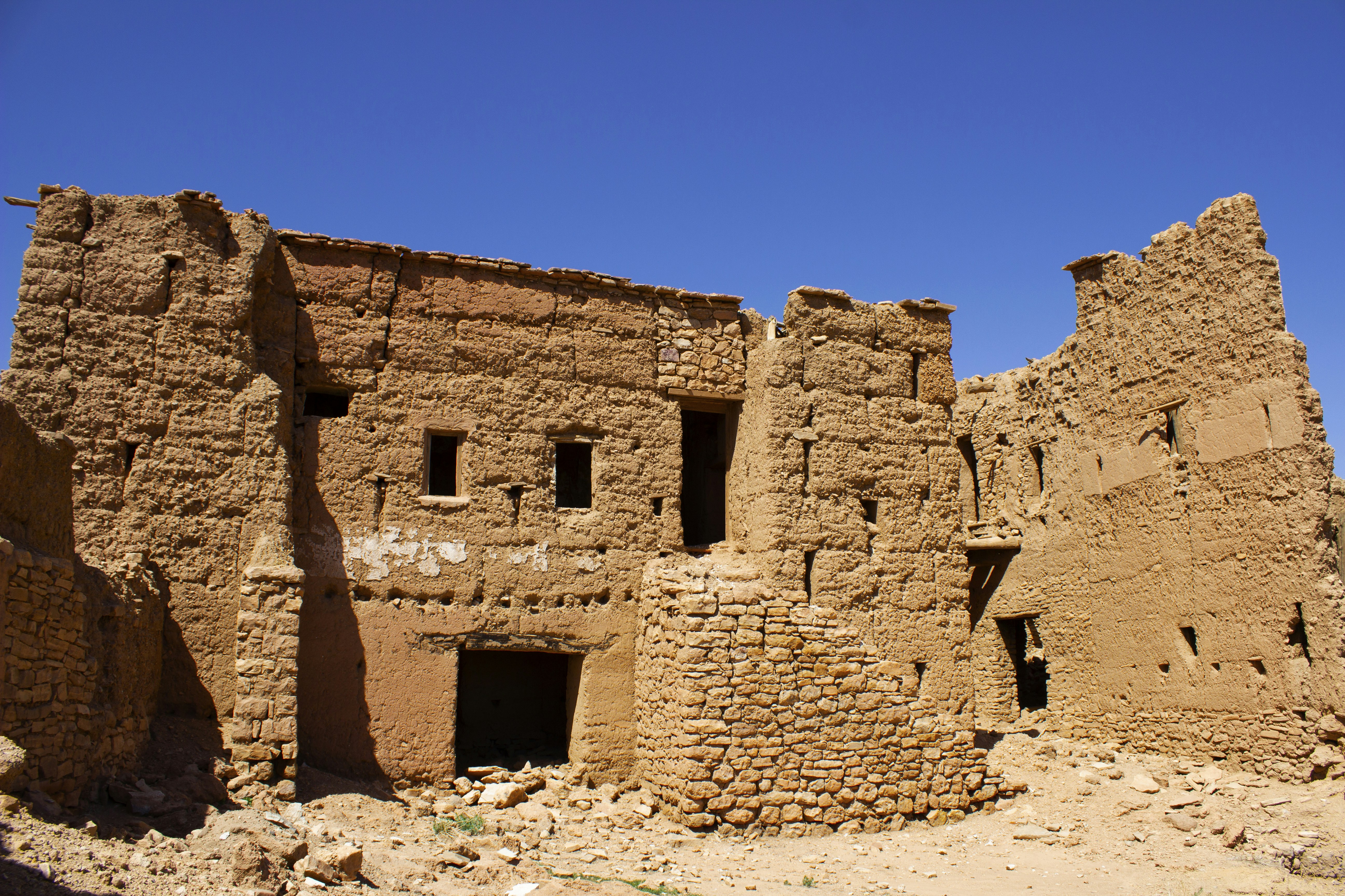 Ancient adobe structure with crumbling walls under a clear blue desert sky, casting deep shadows on the sandy ground.