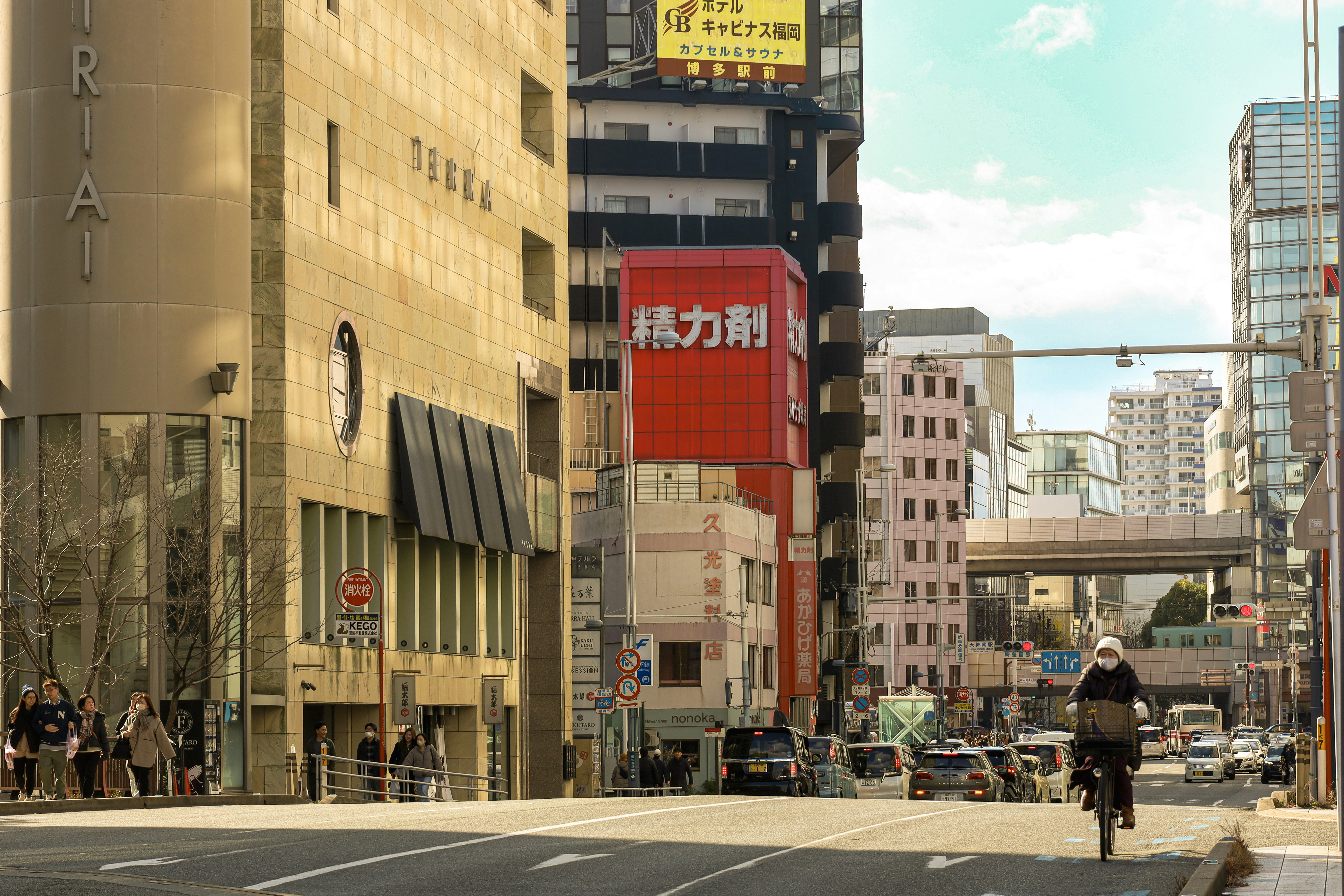 Cyclist rides through a bustling Fukuoka street, framed by modern architecture and vibrant signage under a bright sky.