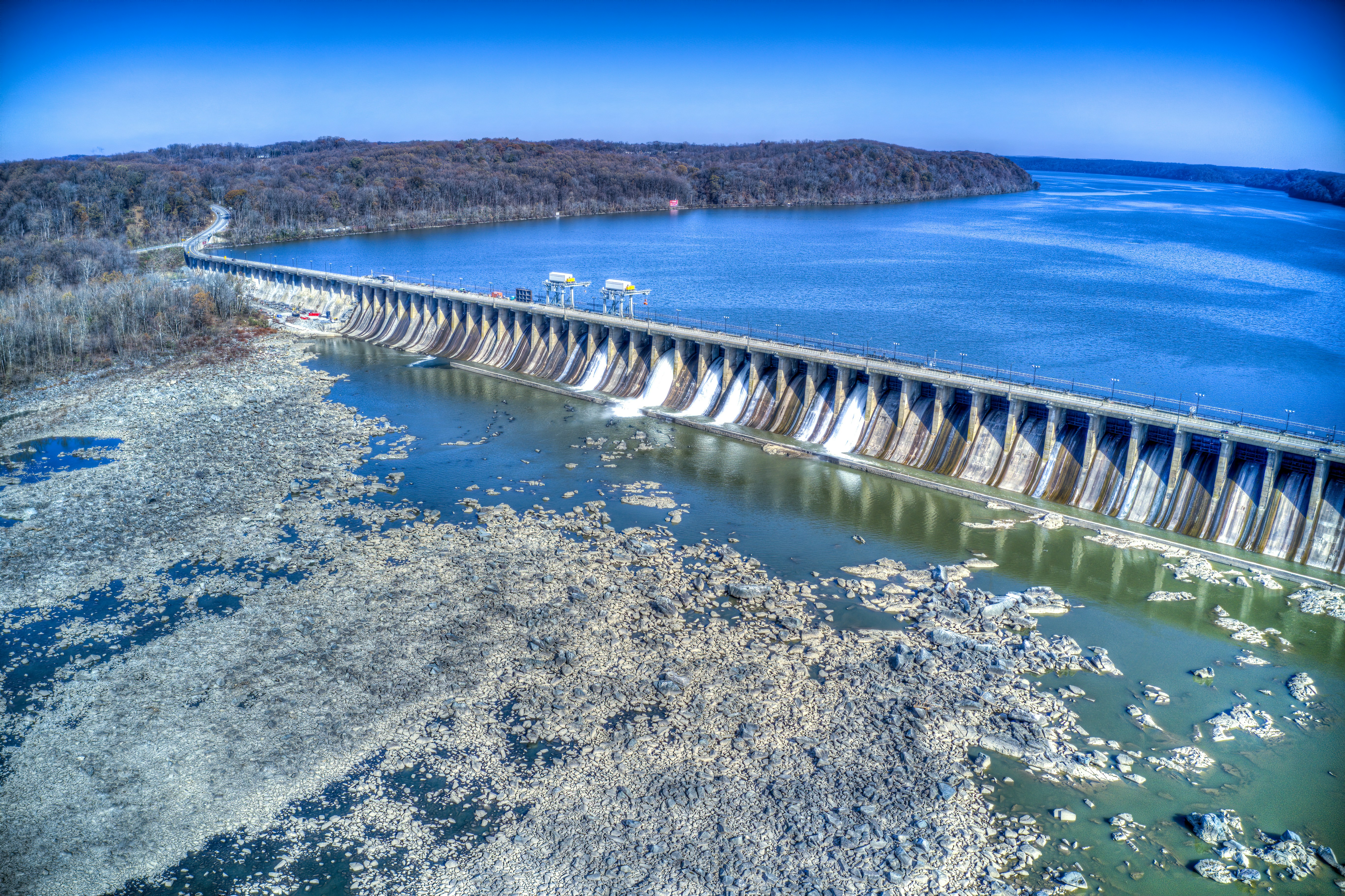 Expansive dam structure spanning a wide river with surrounding rugged landscape under a bright blue sky.