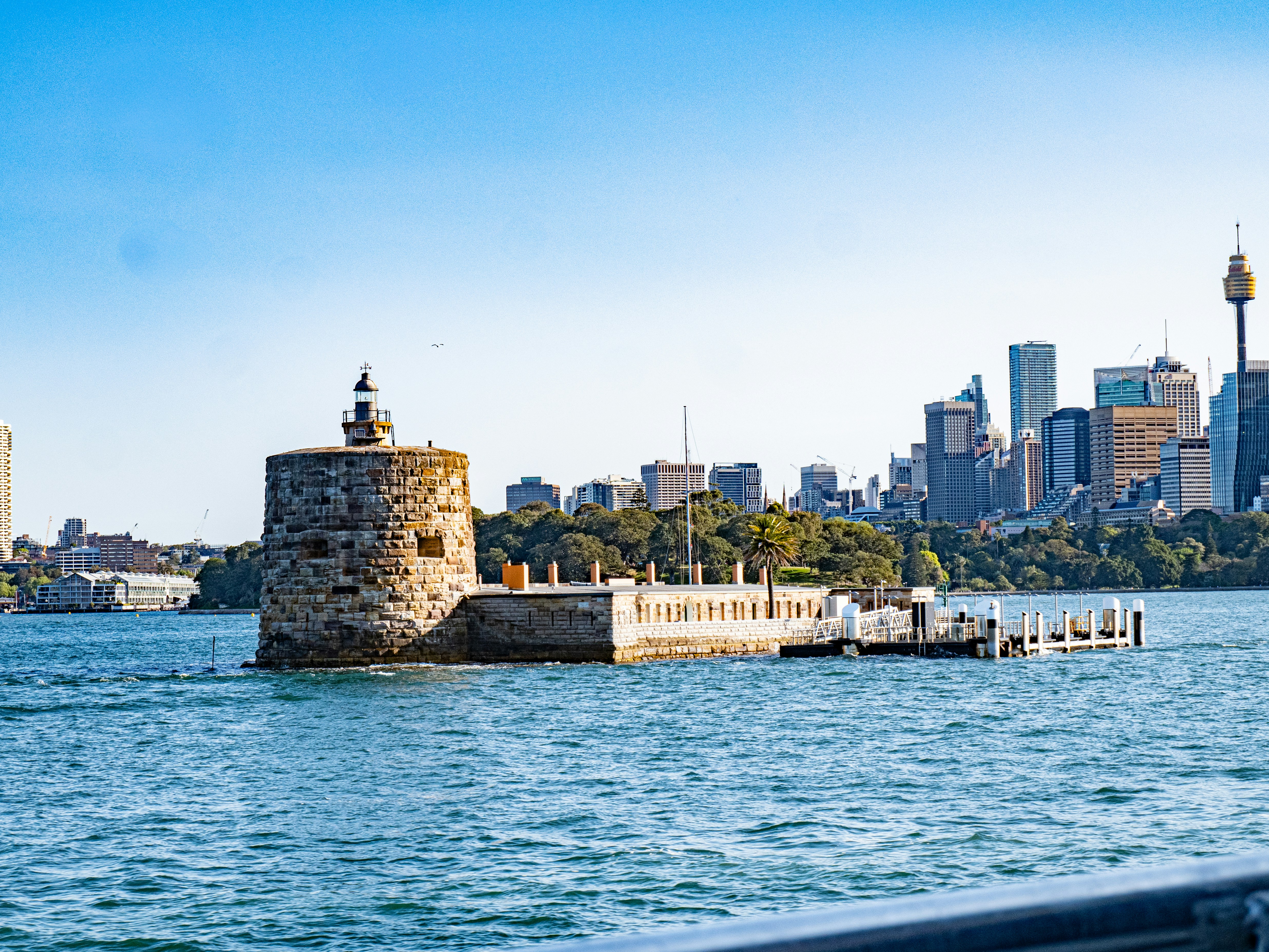 Historic stone lighthouse on a waterfront with a modern city skyline in the background under a clear blue sky.