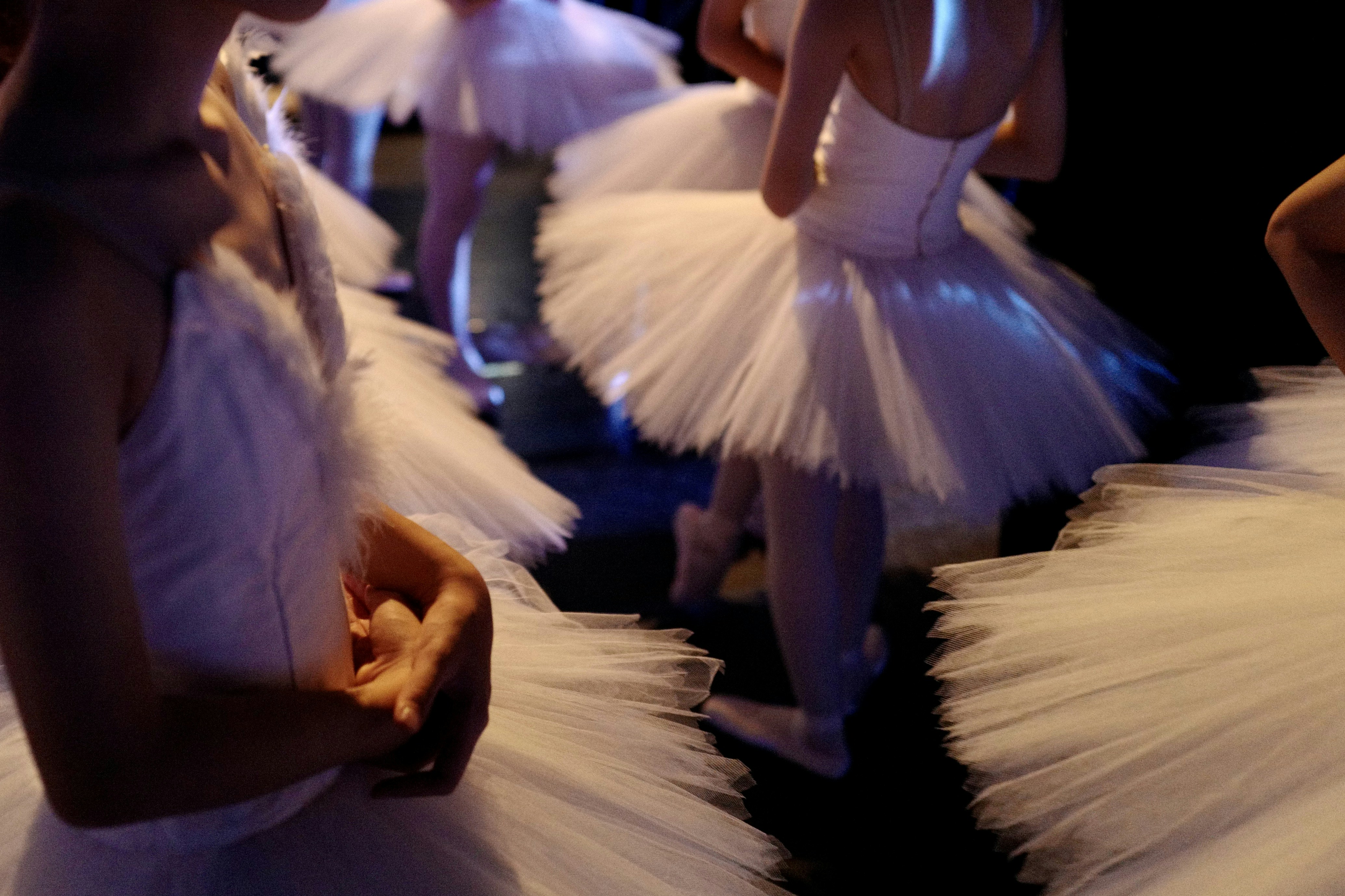 Ballet dancers in white tutus await in dim backstage lighting.
