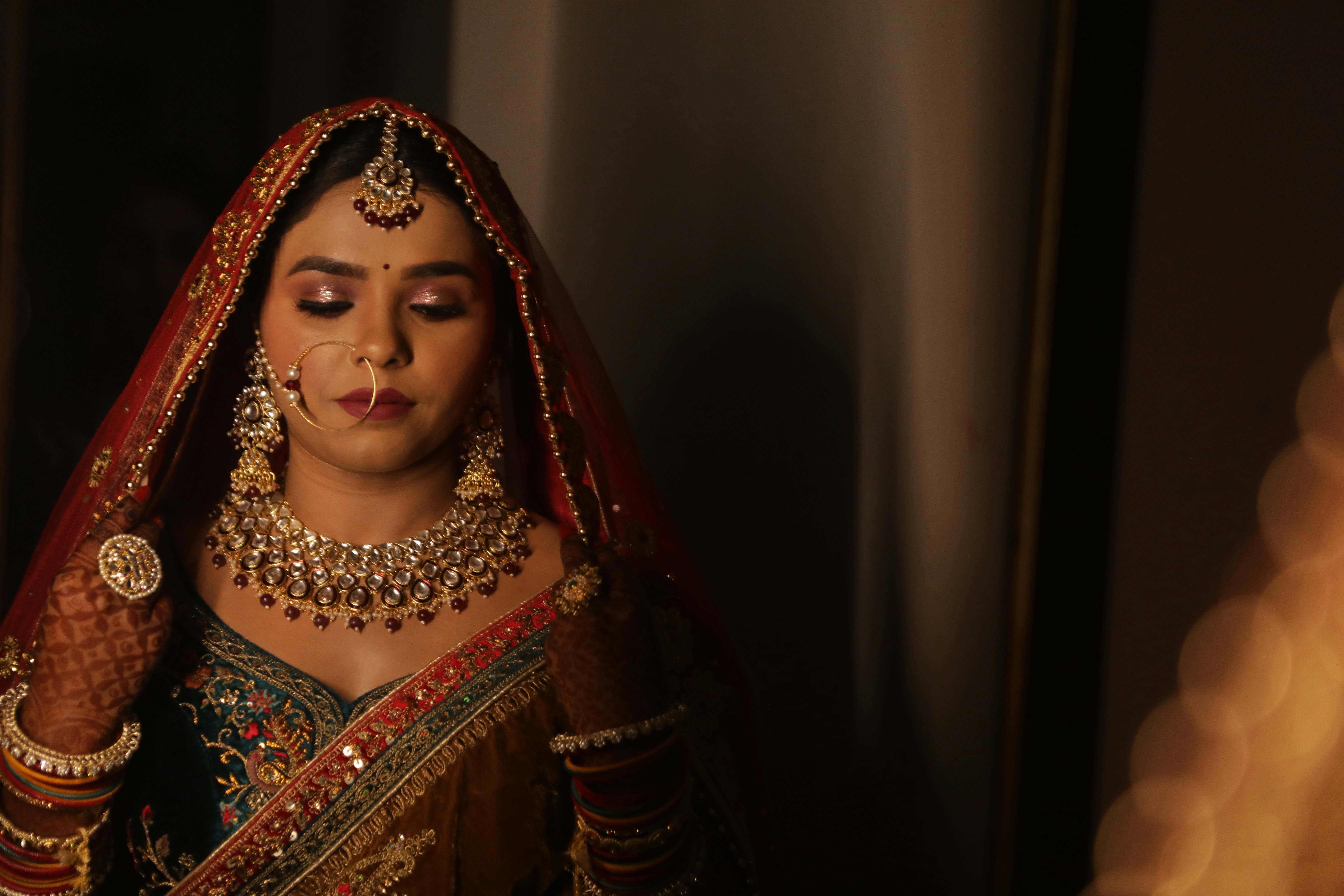 Bride in a red and gold saree adorned with intricate jewelry, bathed in warm light and casting a serene expression.