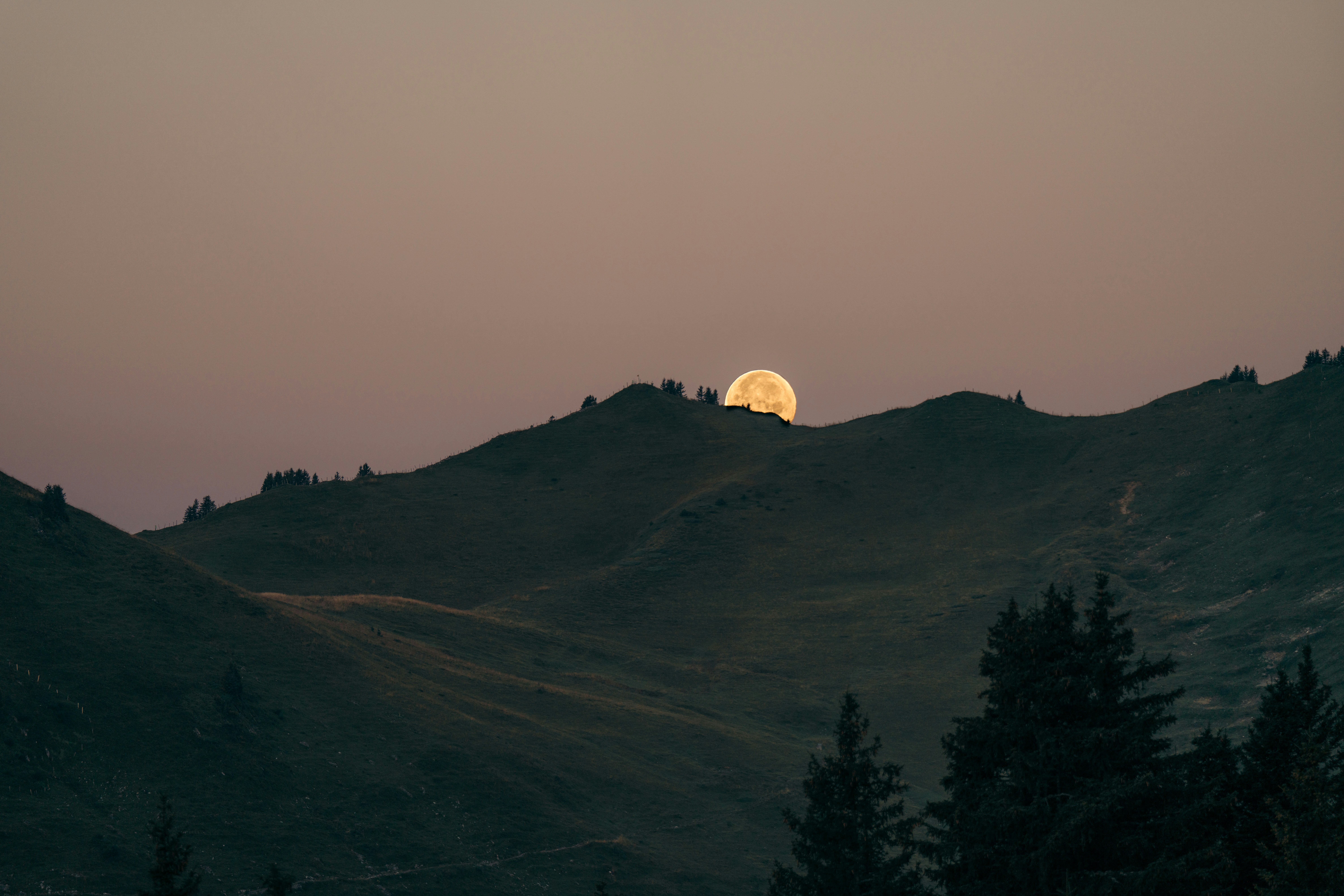 Moon rising above shadowed hills with a soft gradient sky, illuminating the landscape in warm tones.