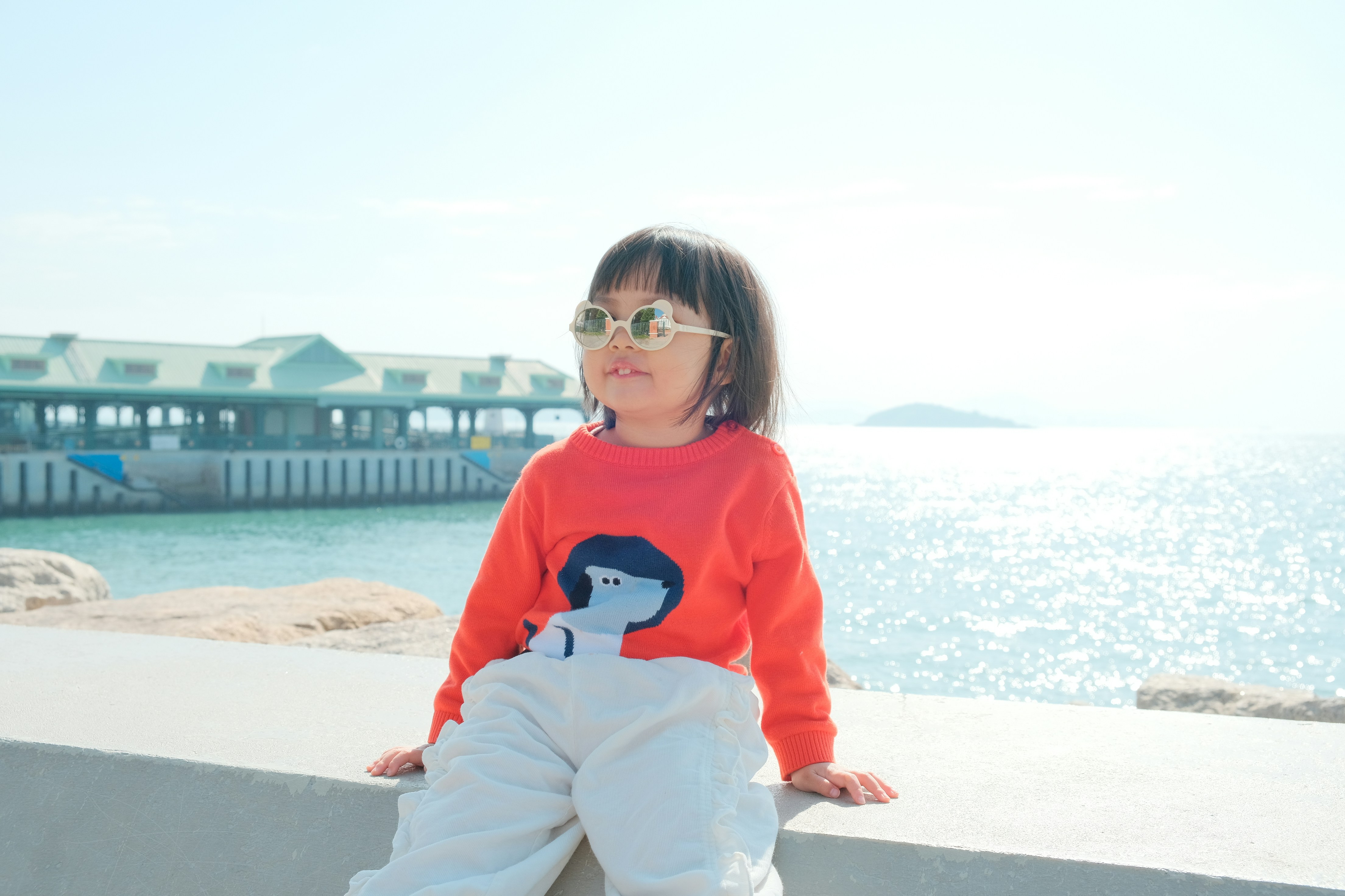 Child in a red sweater sits on a ledge by the ocean, wearing sunglasses with a modern pier in the background.
