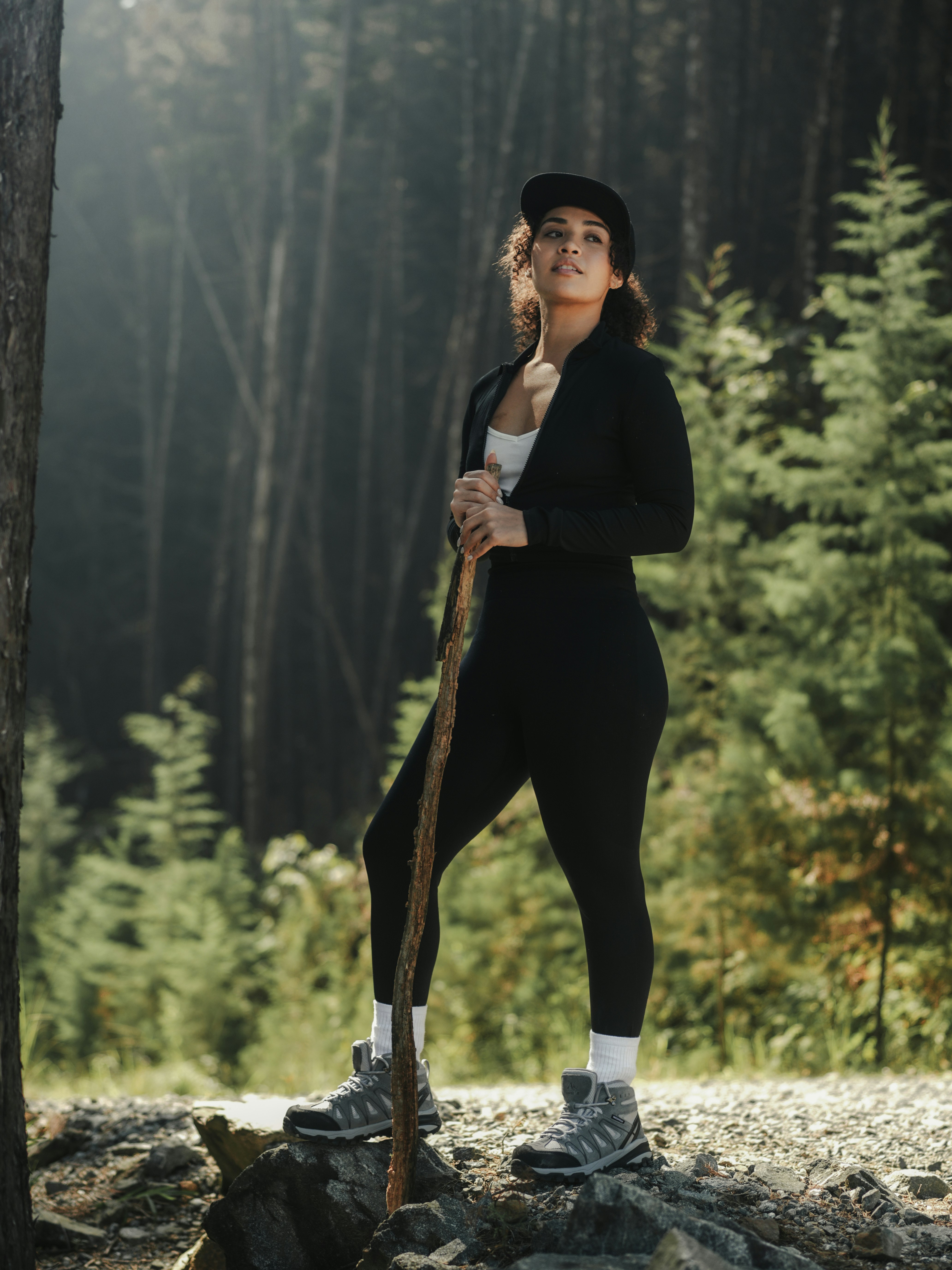 Photograph of a woman in black athletic wear standing on a rocky forest trail, gripping a long staff.