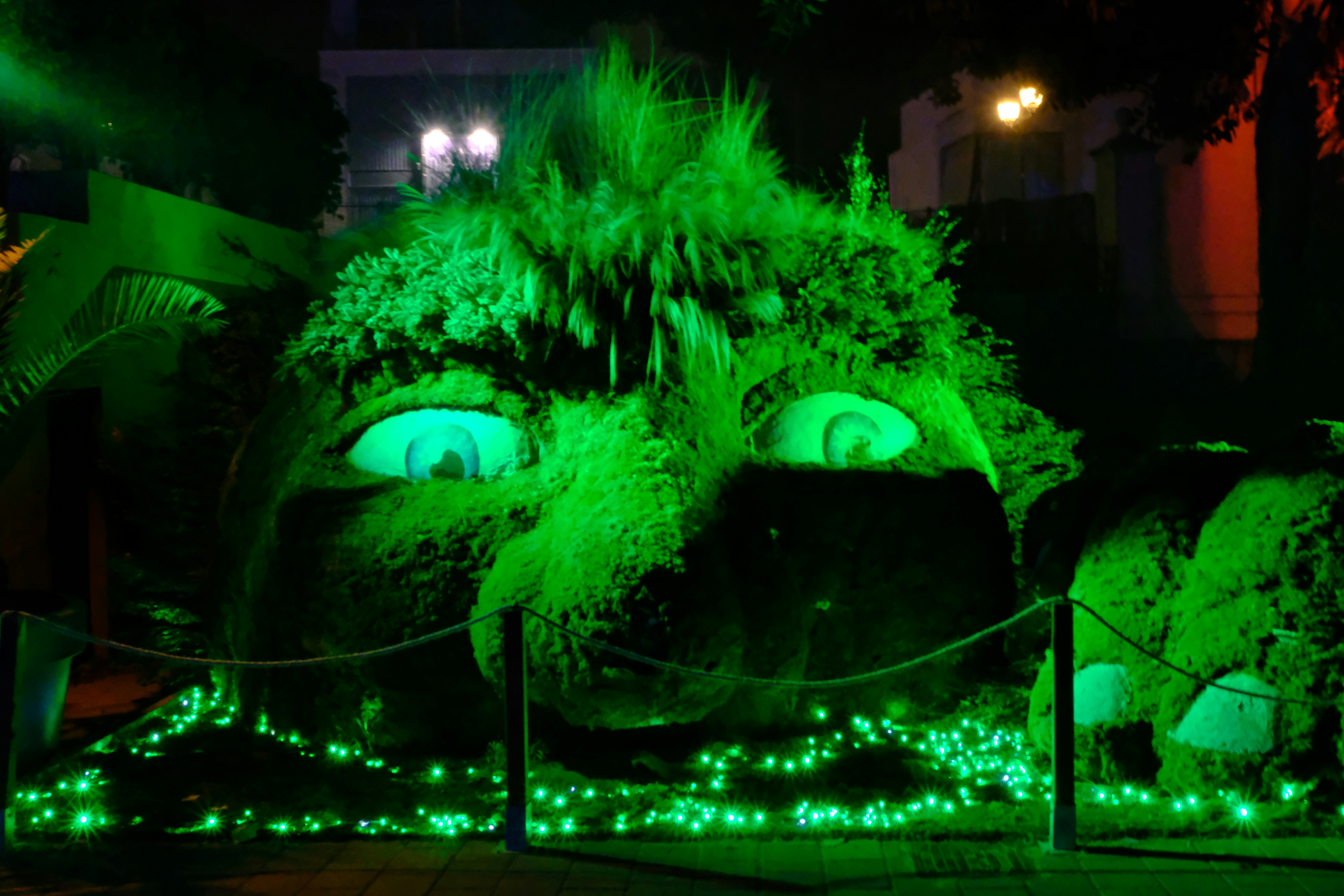 Giant moss-covered sculpture illuminated with vibrant green lights in a nighttime garden setting.