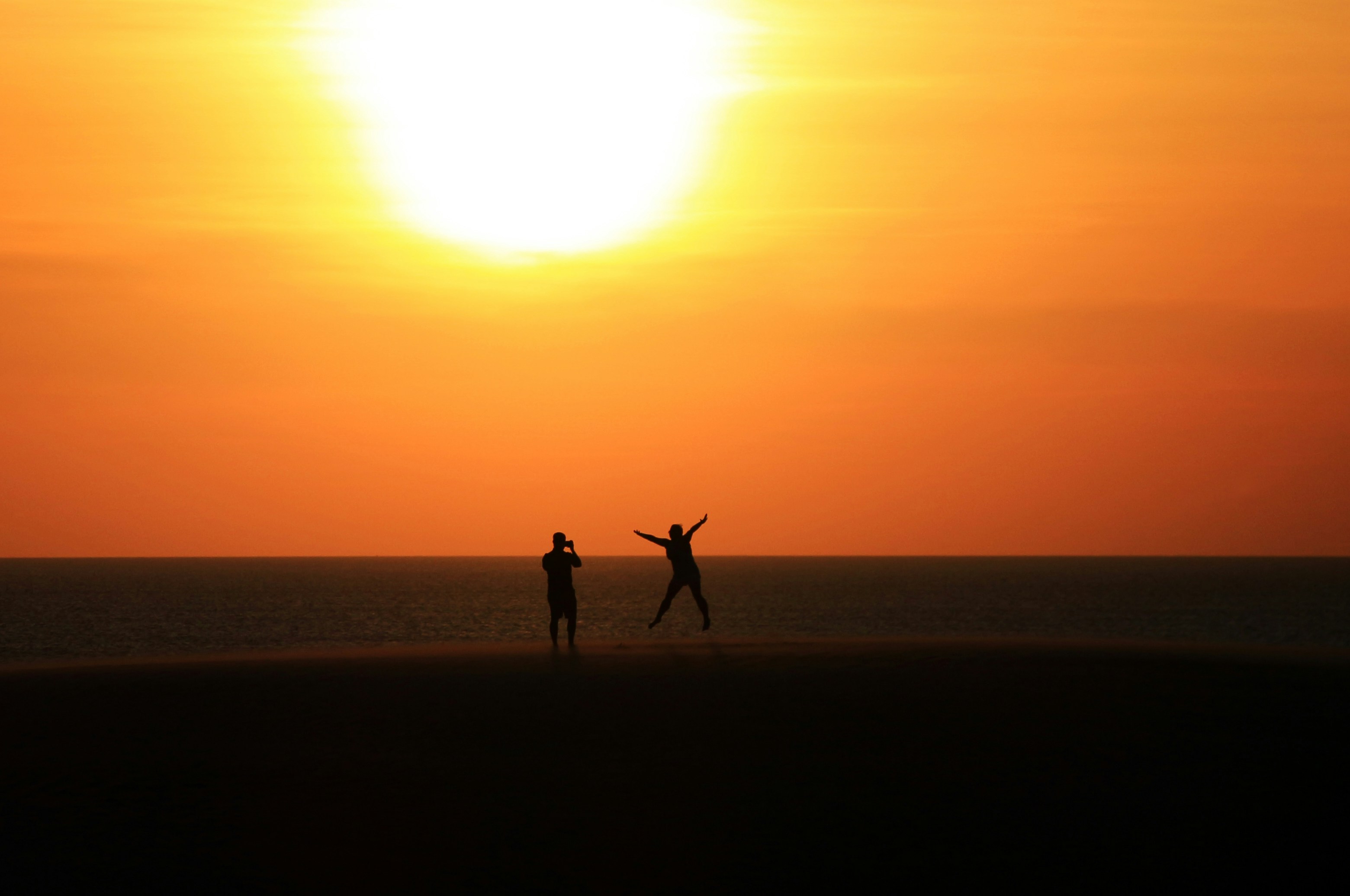 Silhouettes of two figures against a vivid sunset over the ocean, with one figure capturing the moment on camera.