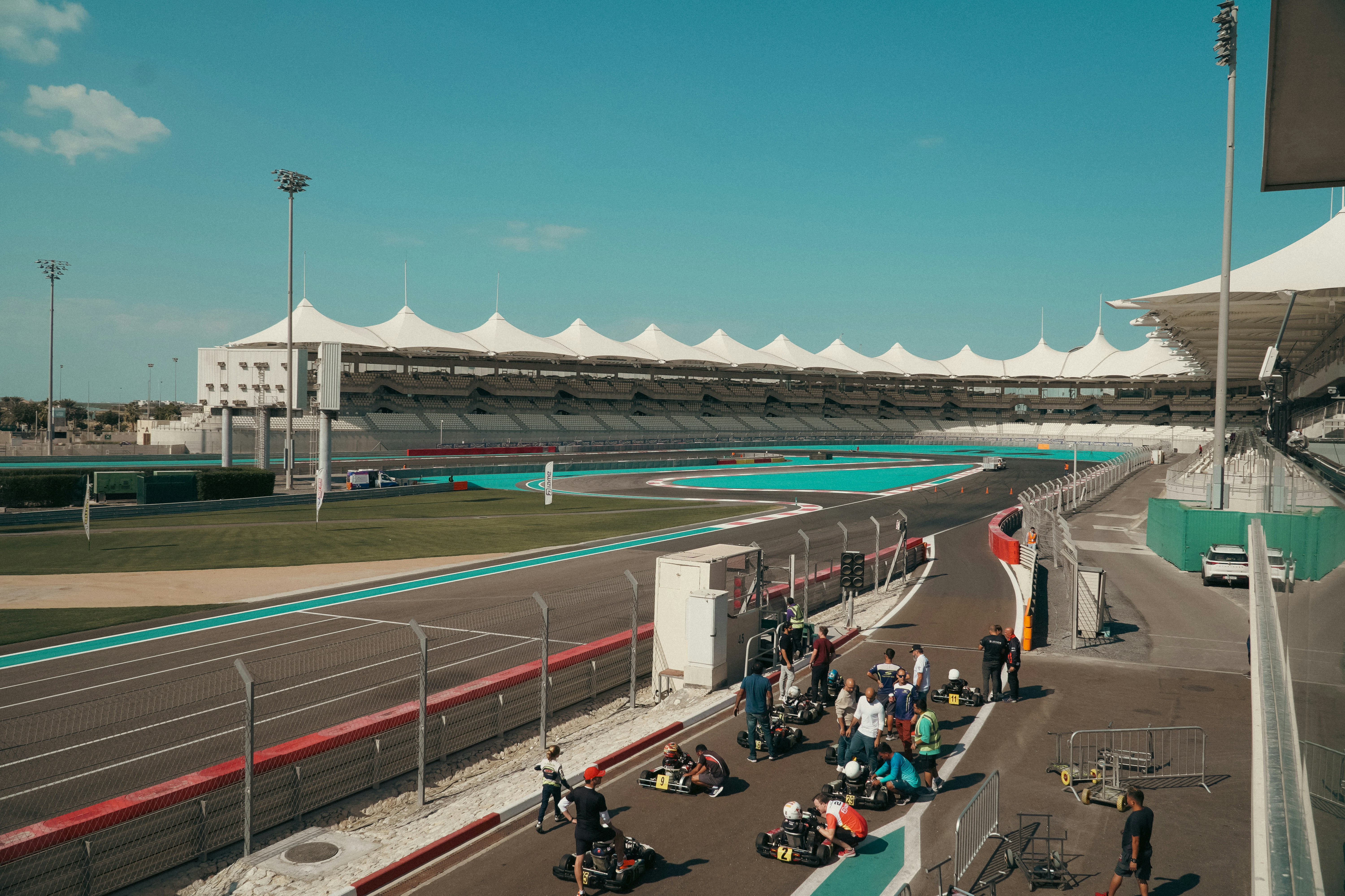 Go-kart racers gather on a vibrant turquoise and asphalt track under a clear blue sky, flanked by white canopies and a grandstand.