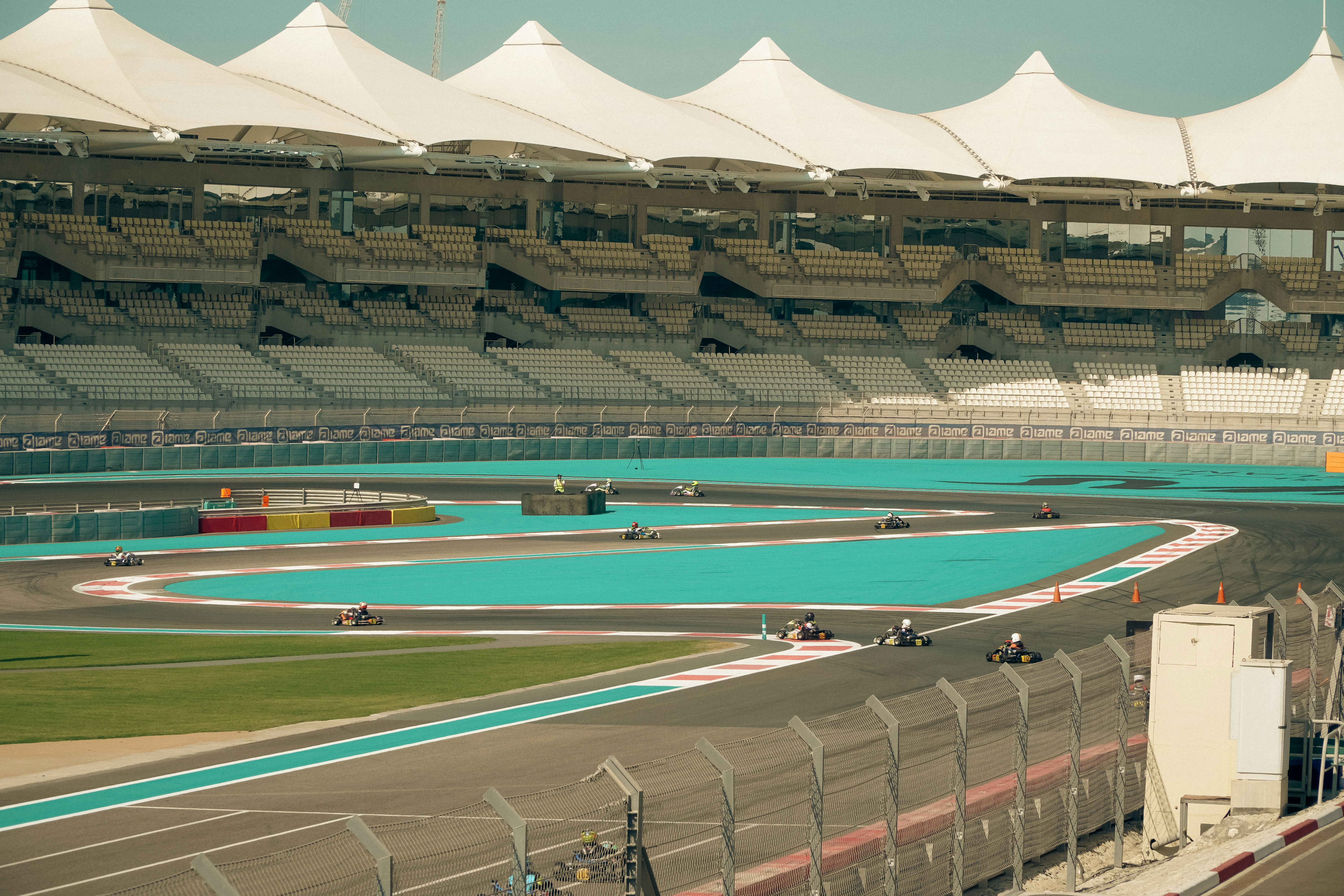 Go-karts racing around a vibrant turquoise track under a grand stadium with white canopies.