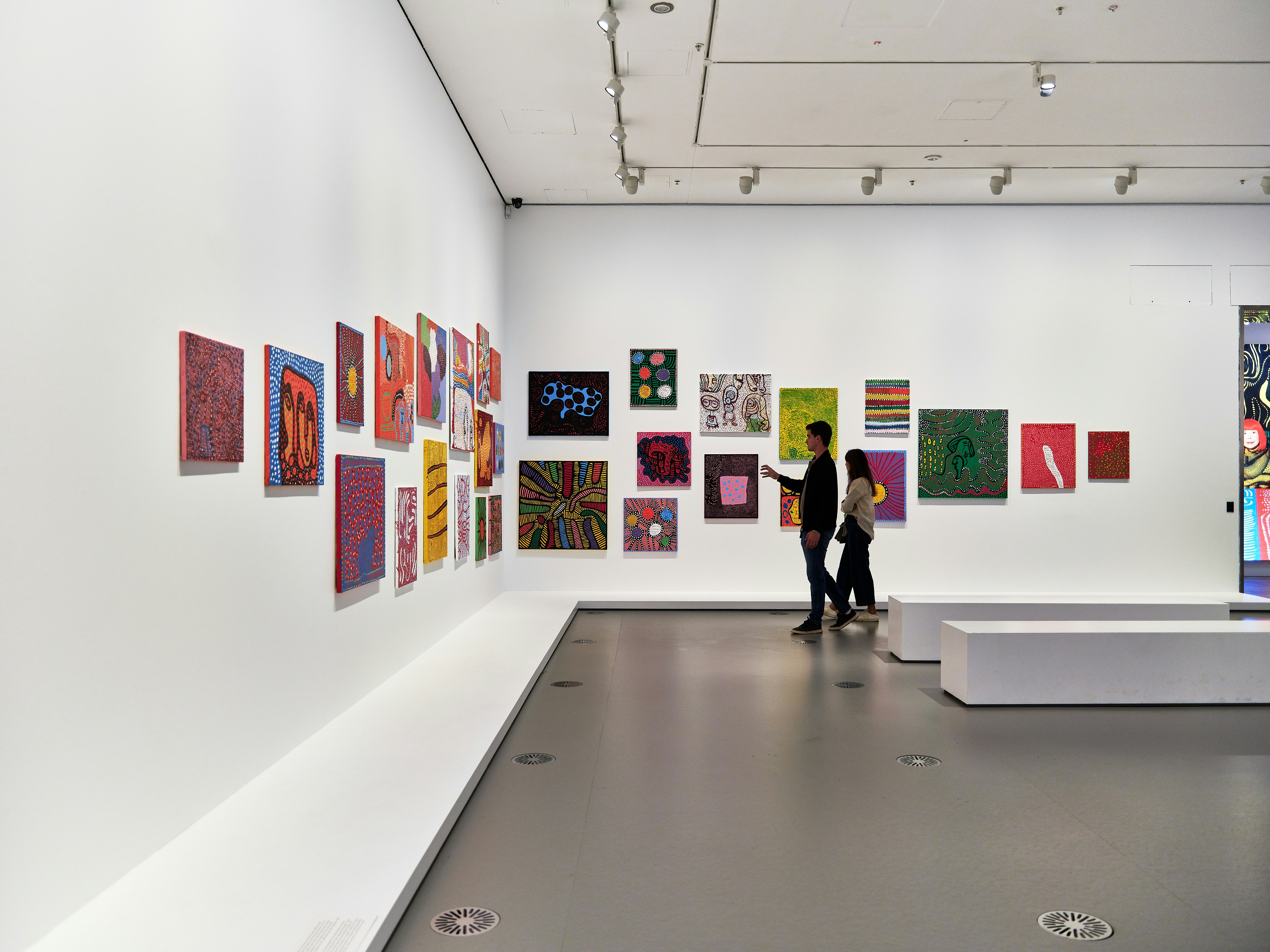 Colorful abstract artworks adorn a white gallery wall as two individuals engage with the vibrant displays.