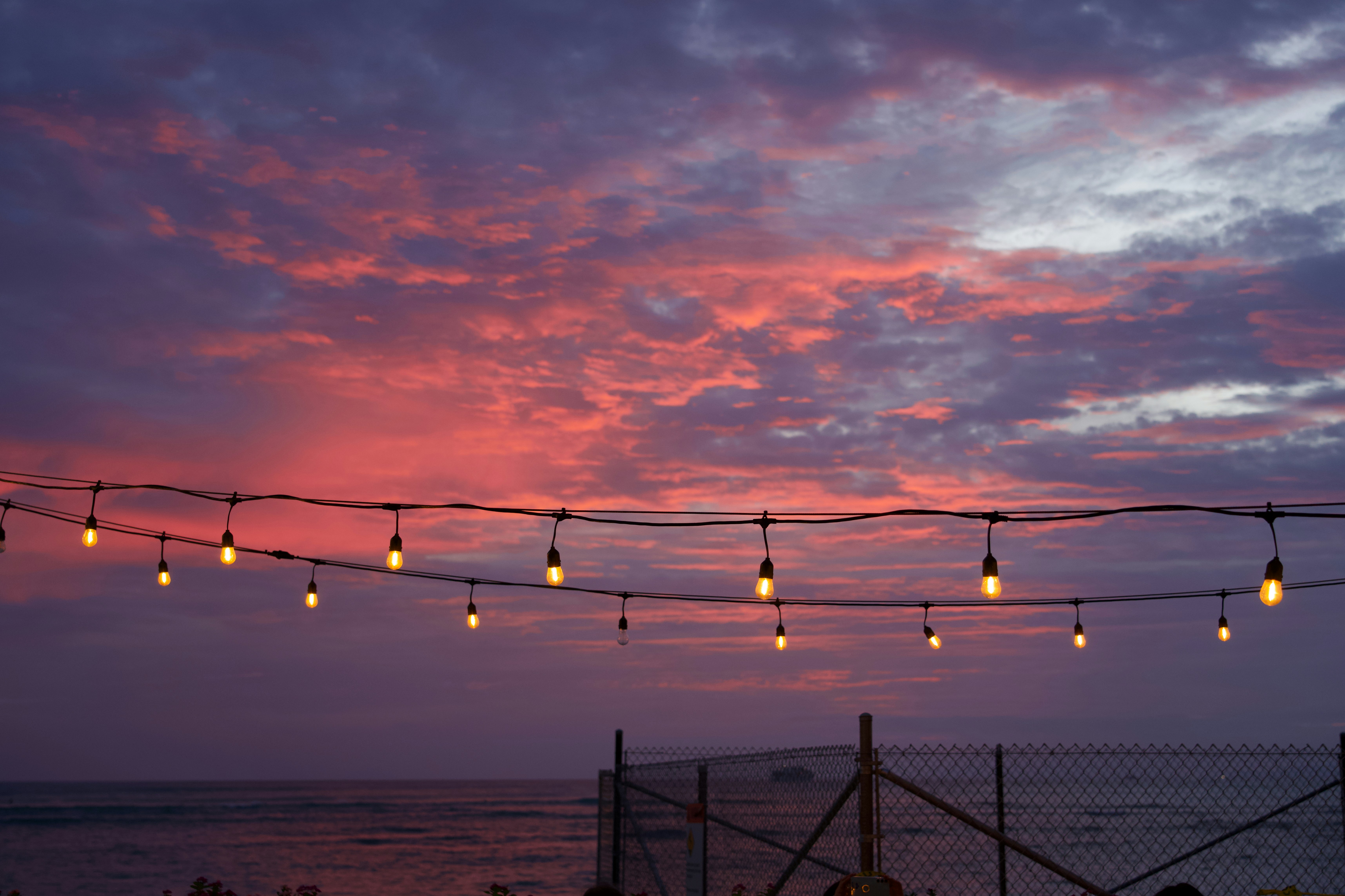 String lights hang above a serene ocean backdrop at sunset, with vibrant pink and purple clouds in the sky.