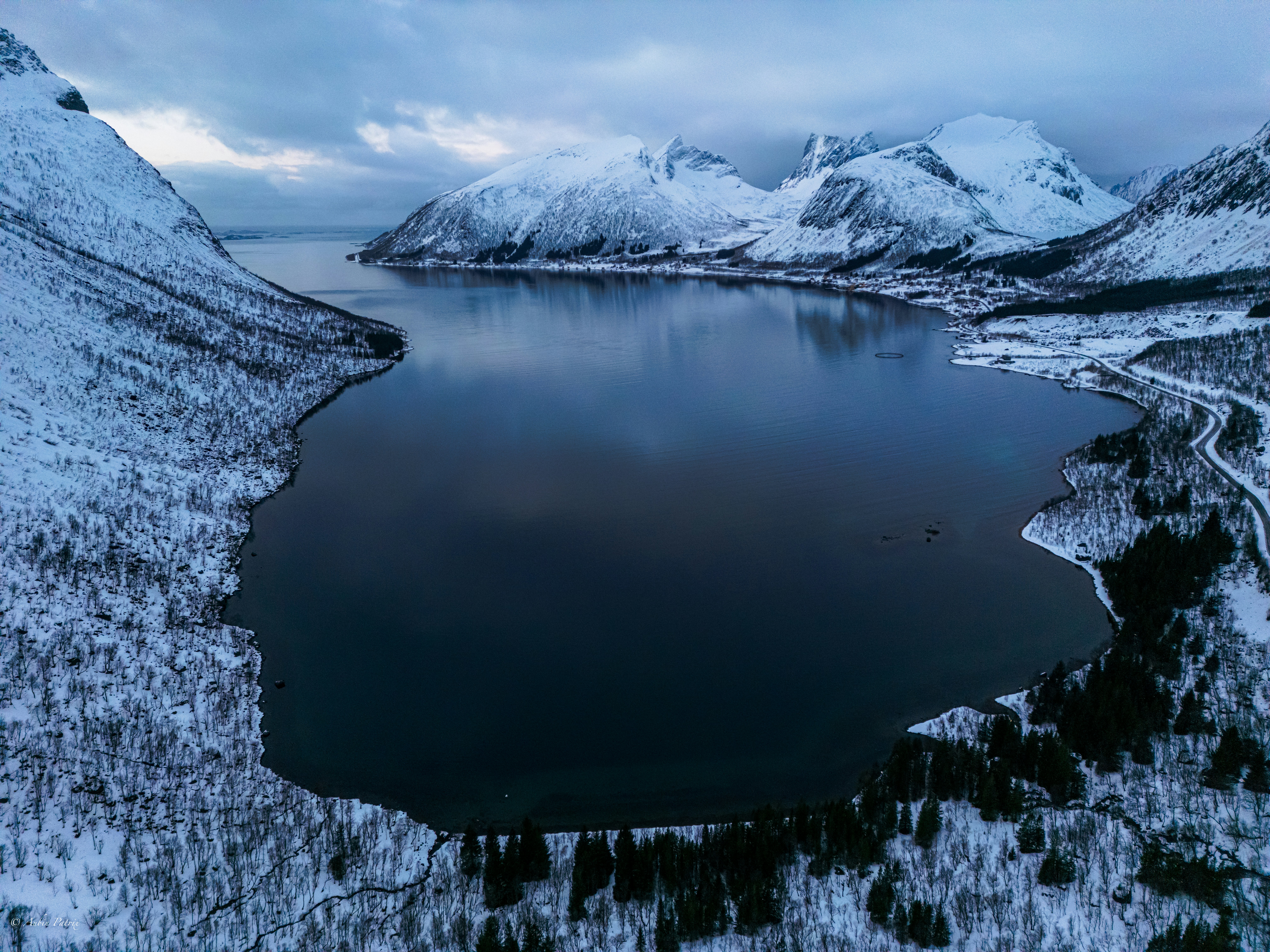 Snow-covered mountains encircle a tranquil fjord under a moody sky.