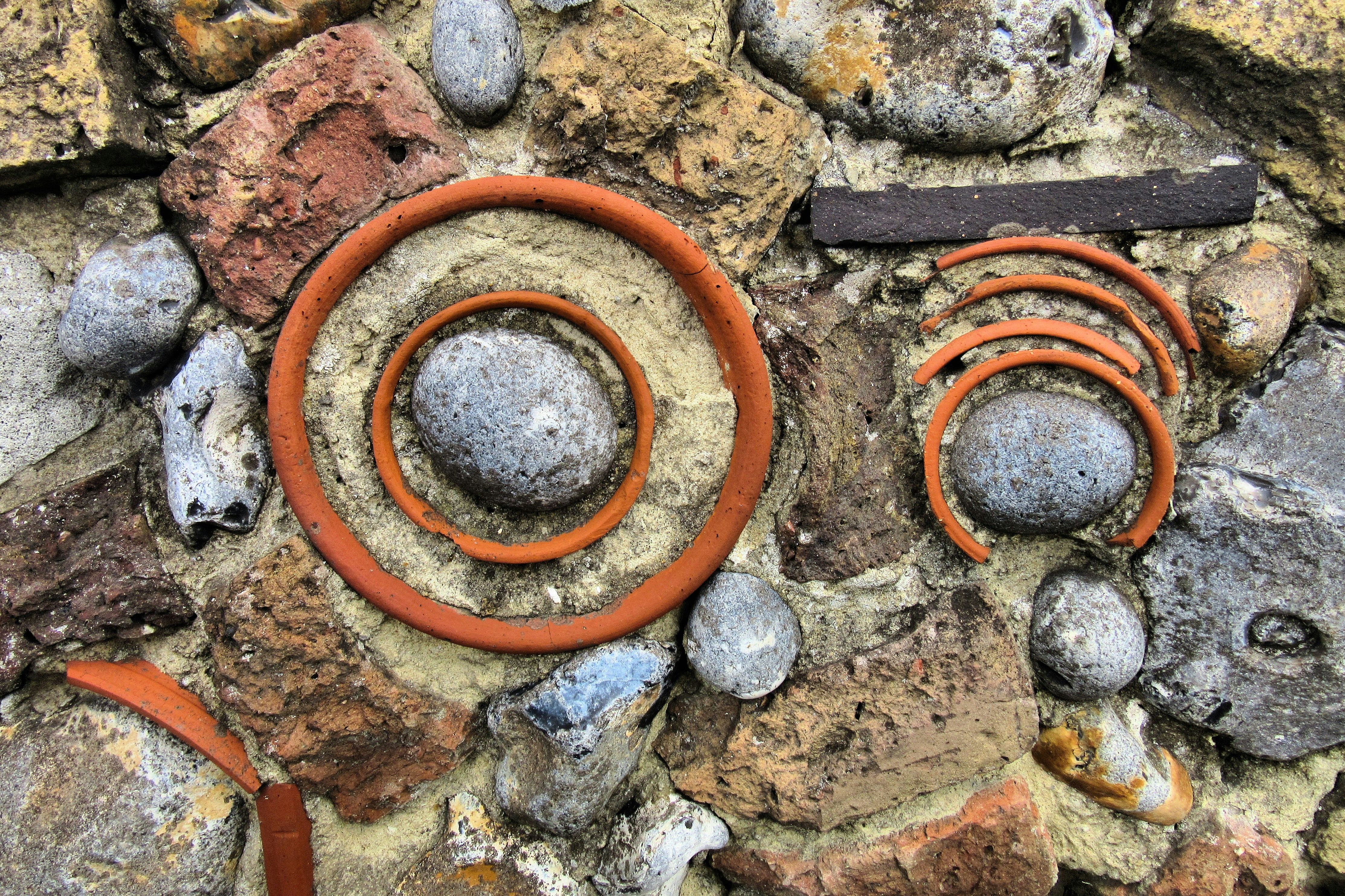 Intricate arrangement of stones and clay circles embedded in a textured wall, showcasing a blend of natural and crafted elements.