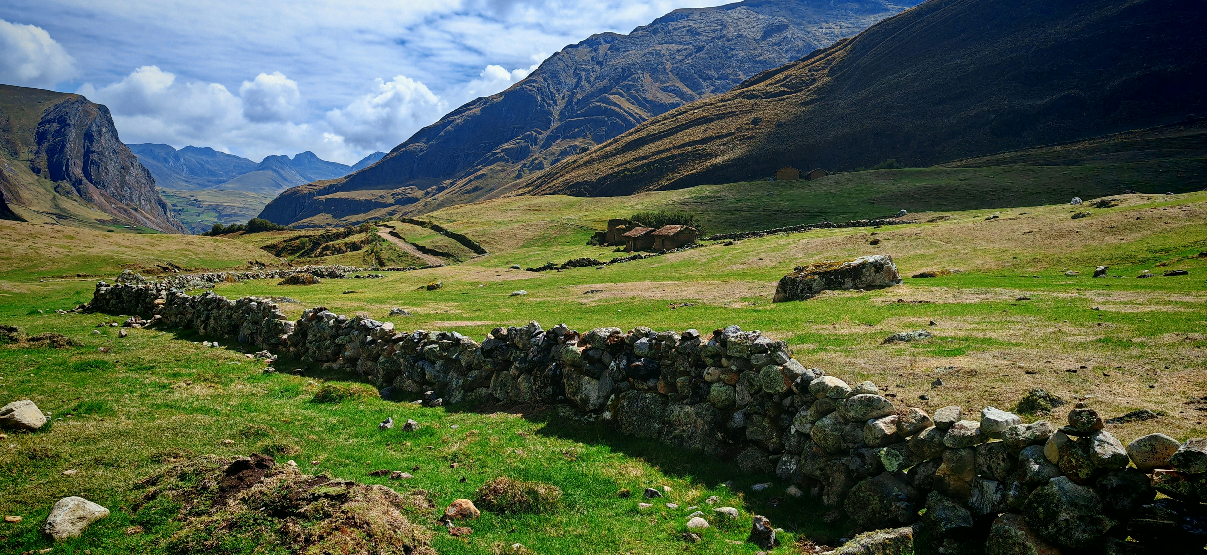 Rolling Highland valley with a weathered dry-stone wall winding toward distant mountains under a bright blue sky. Grass, rocks, and earth tones emphasize texture and rural landscape.