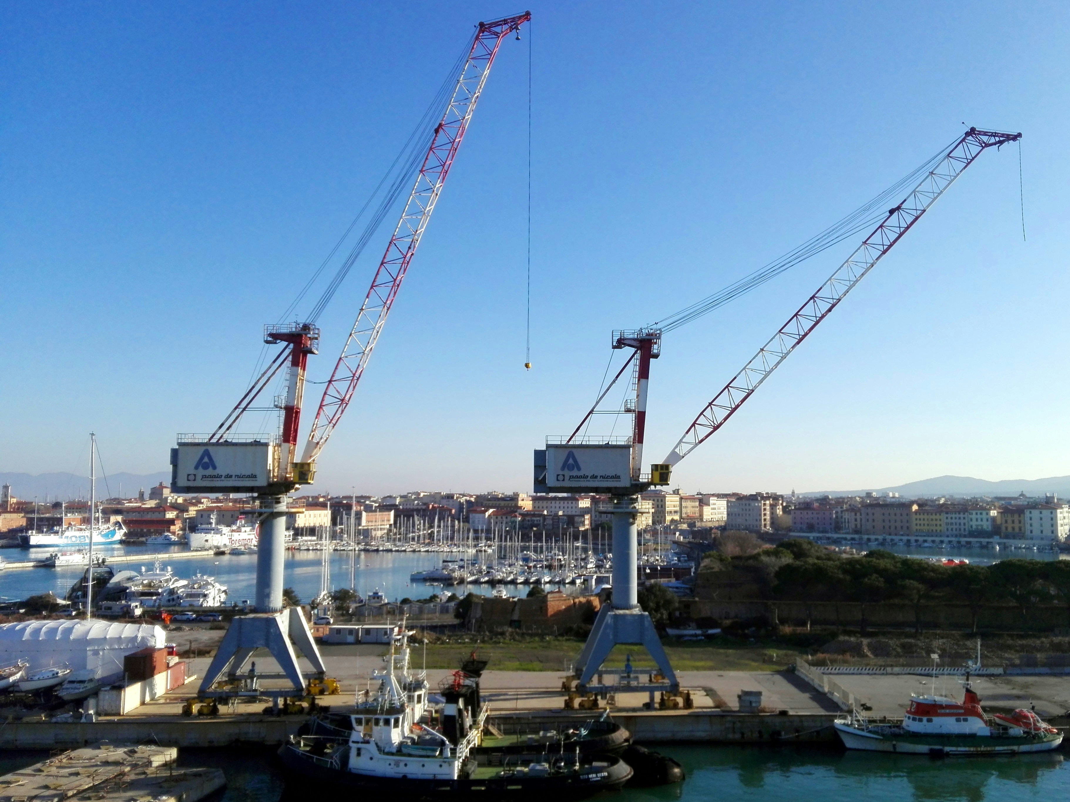 Two red and white cranes tower over a bustling harbor with boats and a distant cityscape under a clear blue sky.
