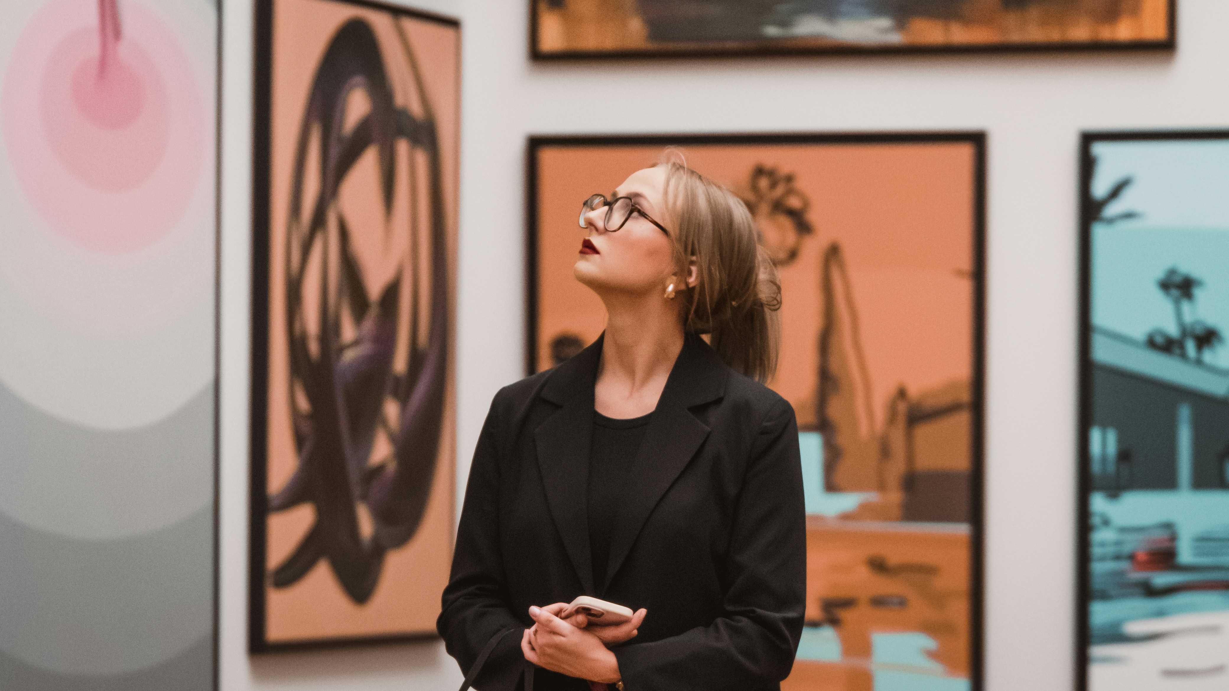 Woman in black blazer gazing at abstract paintings in an art gallery, surrounded by earthy tones and pastel blues.