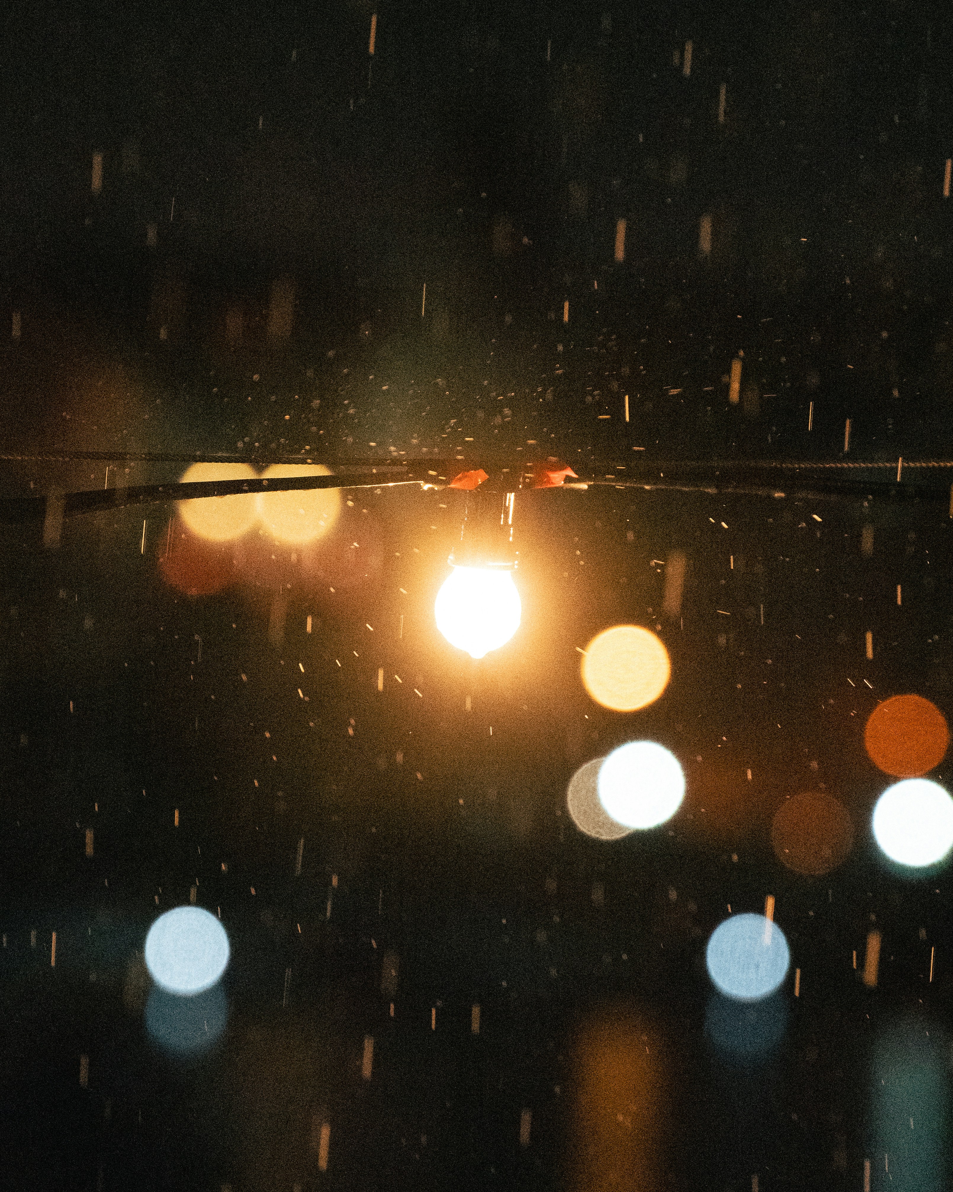 A glowing light bulb suspended in the rain, surrounded by colorful bokeh effects from distant lights. The scene captures a moody ambiance of a rainy night.