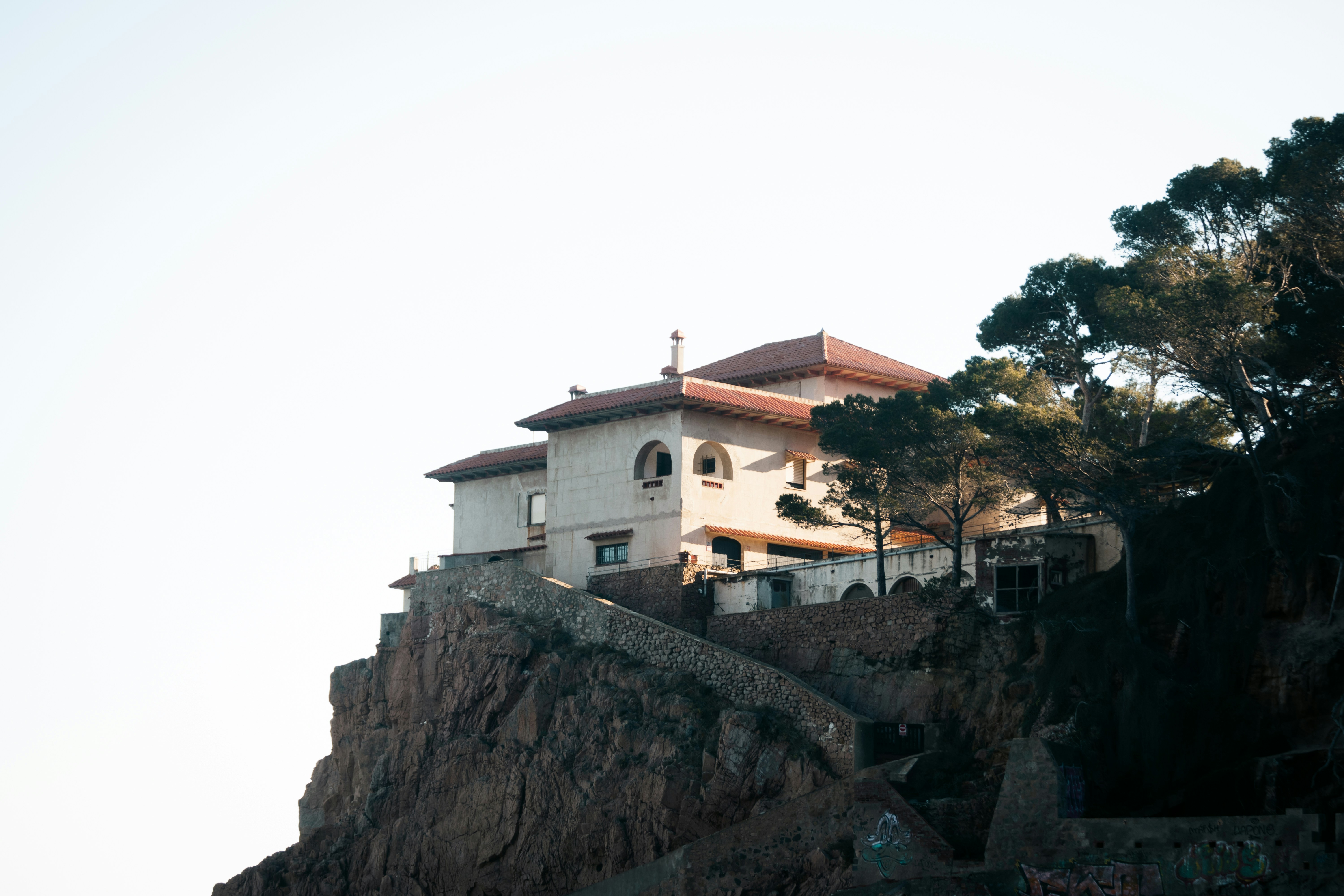 A house with terracotta roof tiles perched on a cliff, framed by trees and illuminated by soft sunlight.