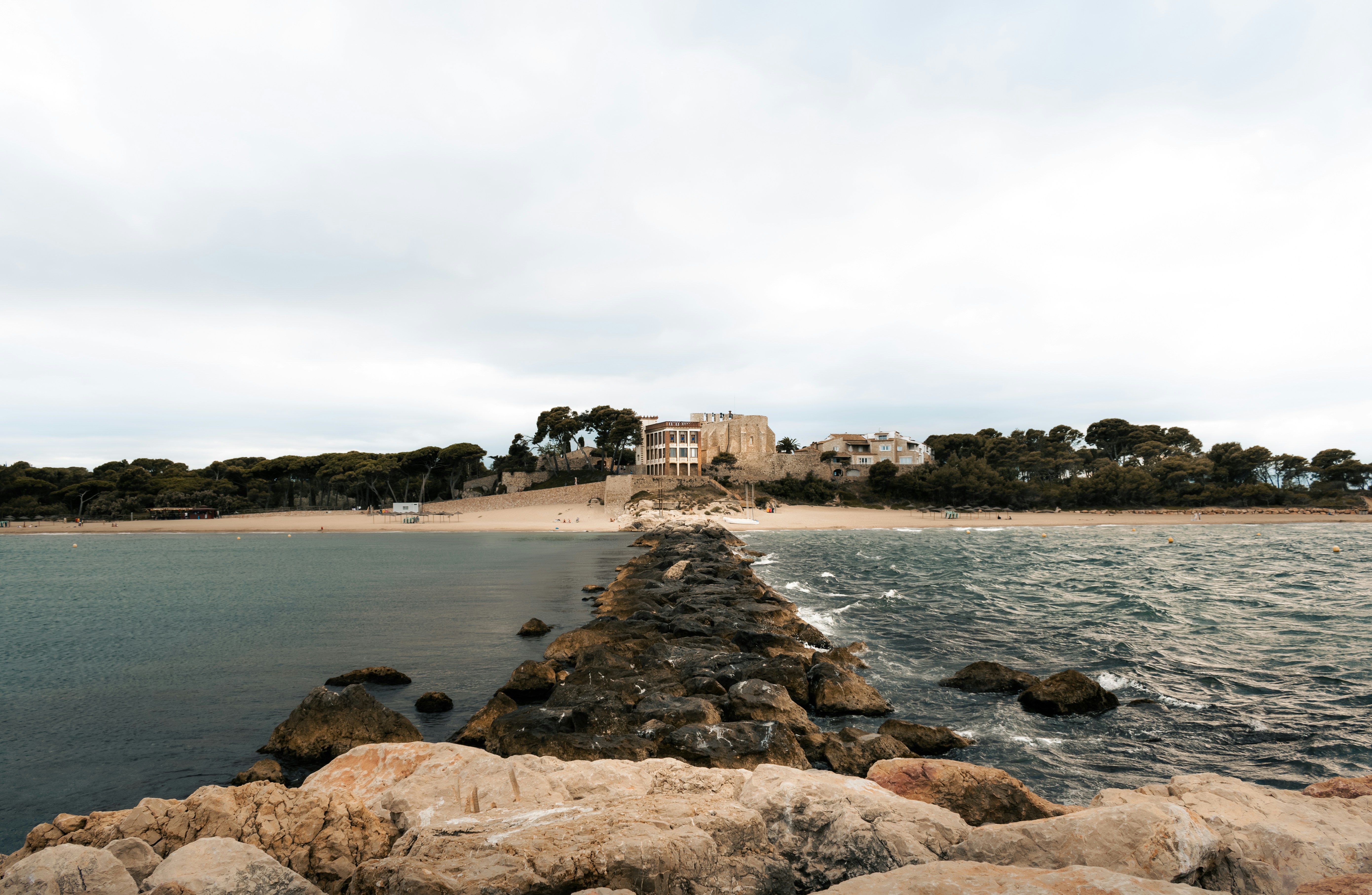 Rocky jetty extends towards a sandy beach lined with greenery under an overcast sky.