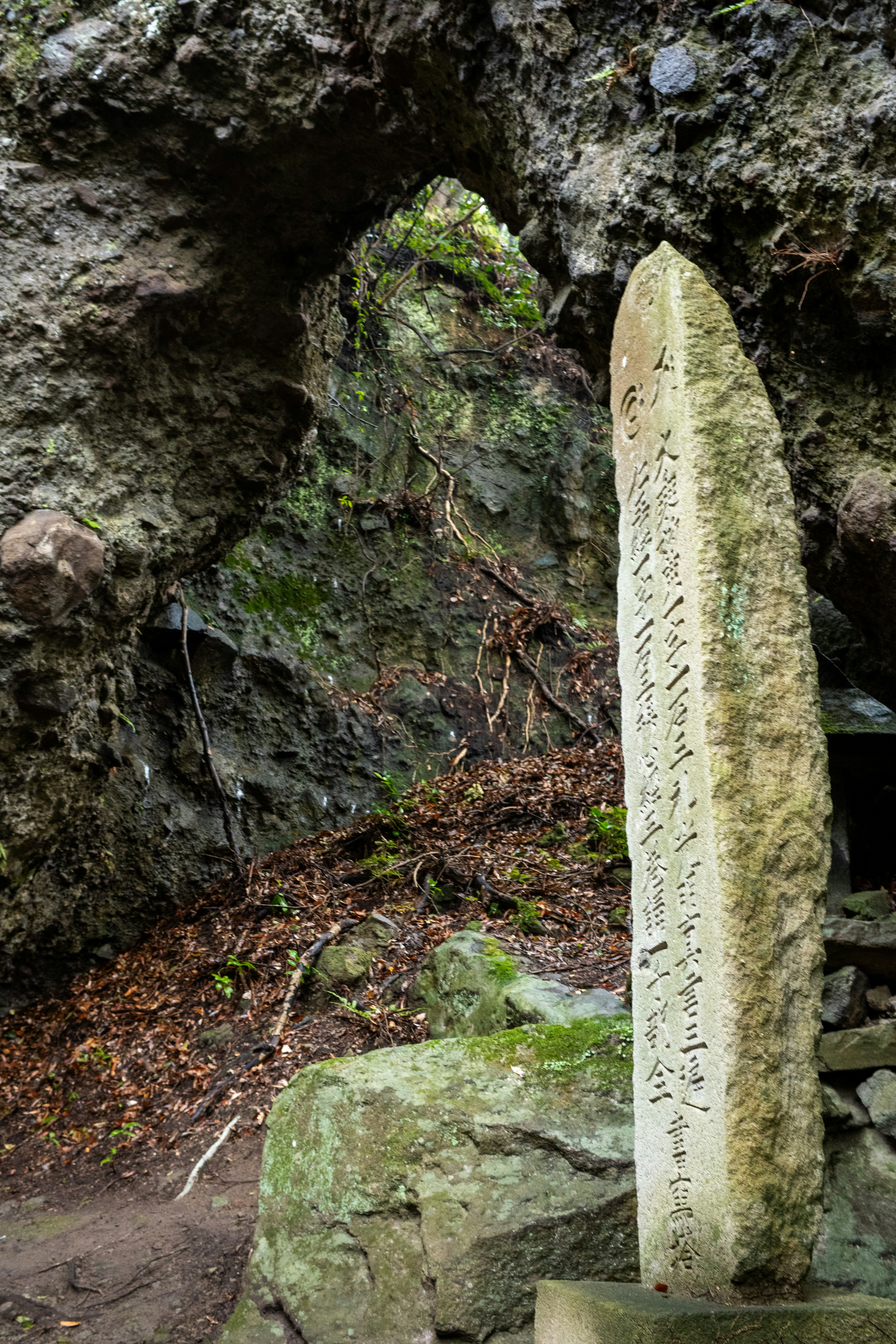 A weathered stone pillar etched with vertical script stands beside a moss-covered rock arch in a dim cave-like alcove.