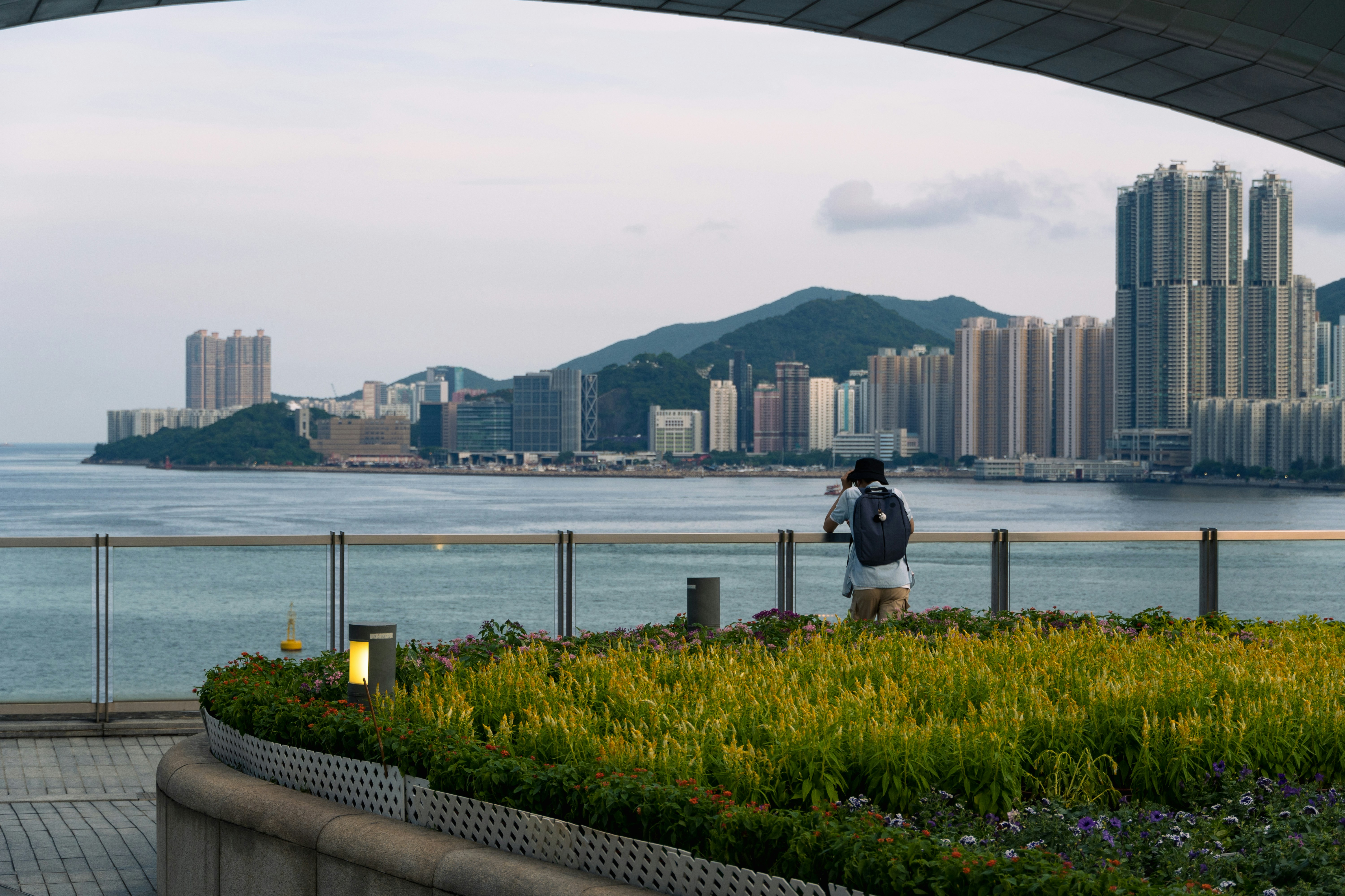 Person with a backpack overlooks a city skyline from a waterfront garden under a wide architectural archway.