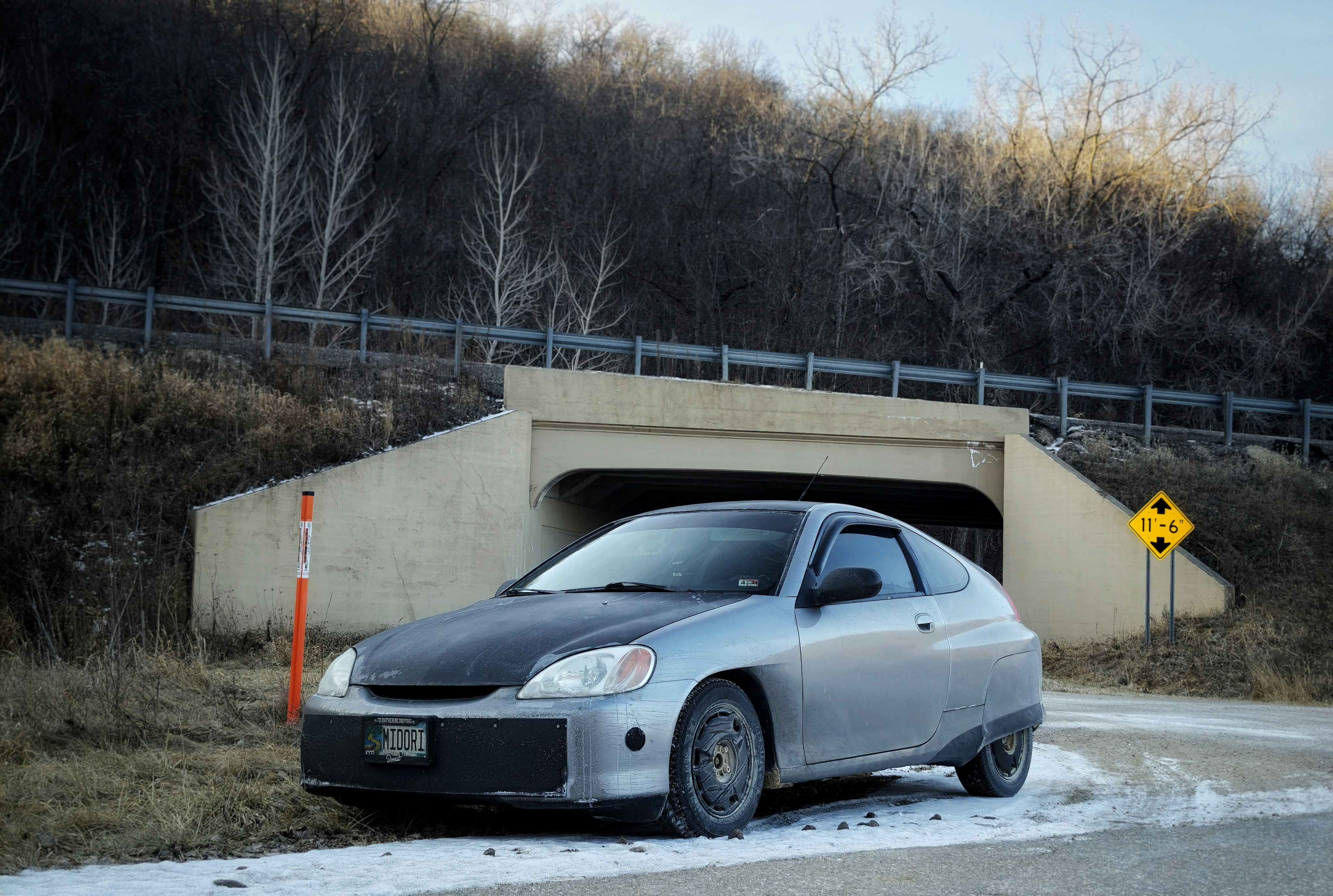 Silver car parked beside a snow-dusted road near an underpass, with barren trees in the background.