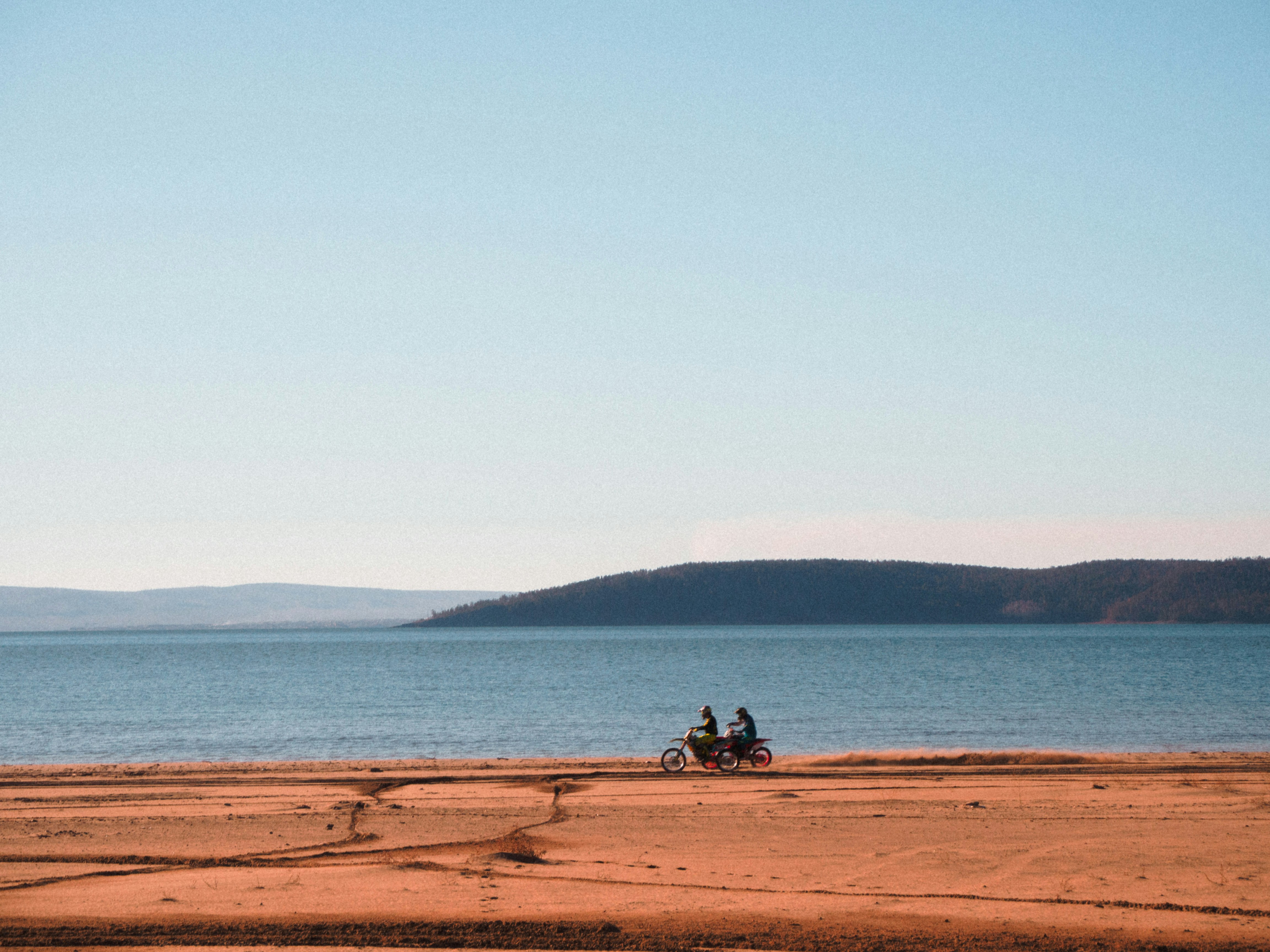 Motorcyclists traverse a sandy shoreline with a calm sea and distant hills under a clear blue sky.