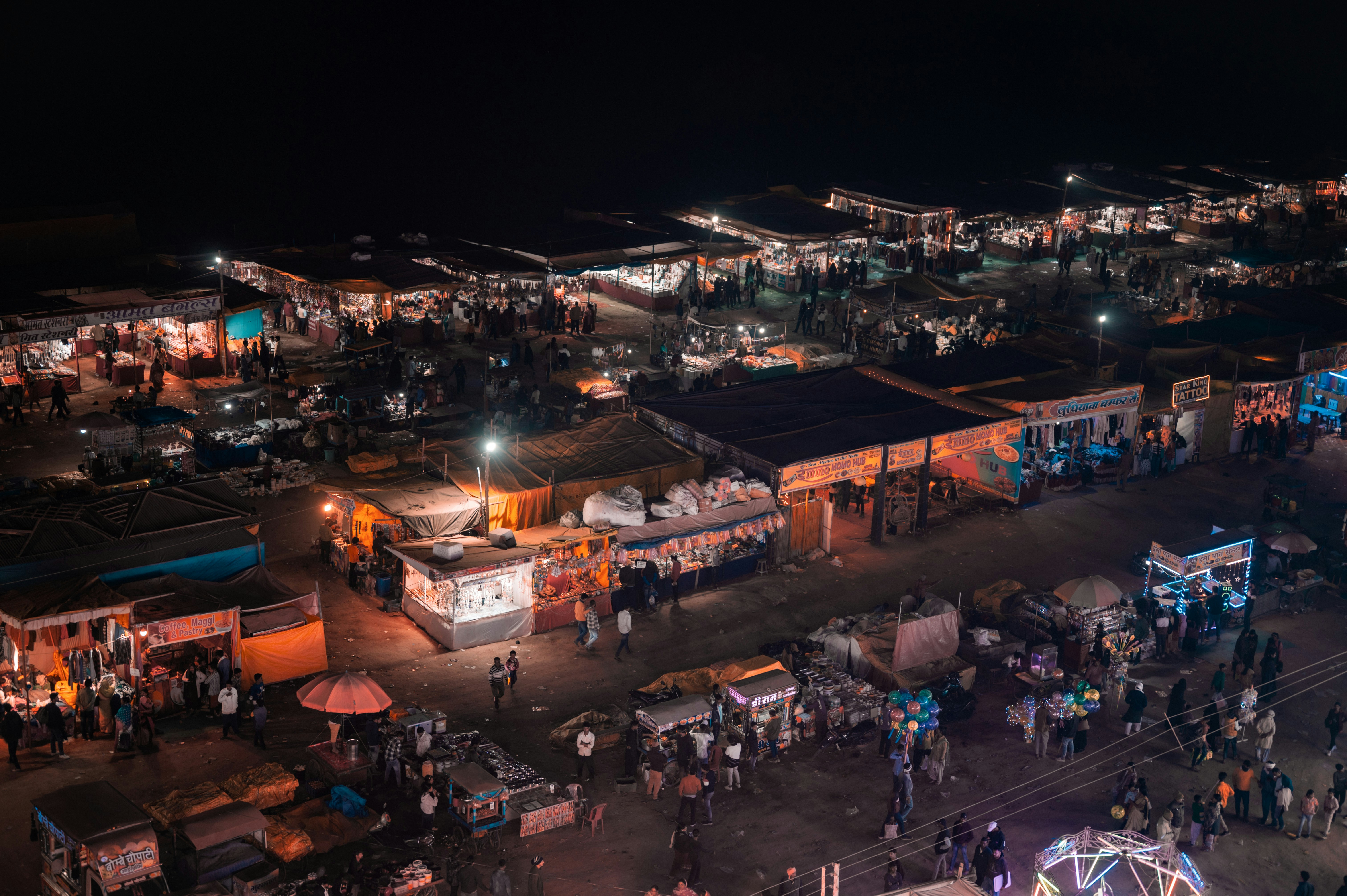 Aerial view of a bustling night market with rows of illuminated stalls and vibrant crowds under the night sky.