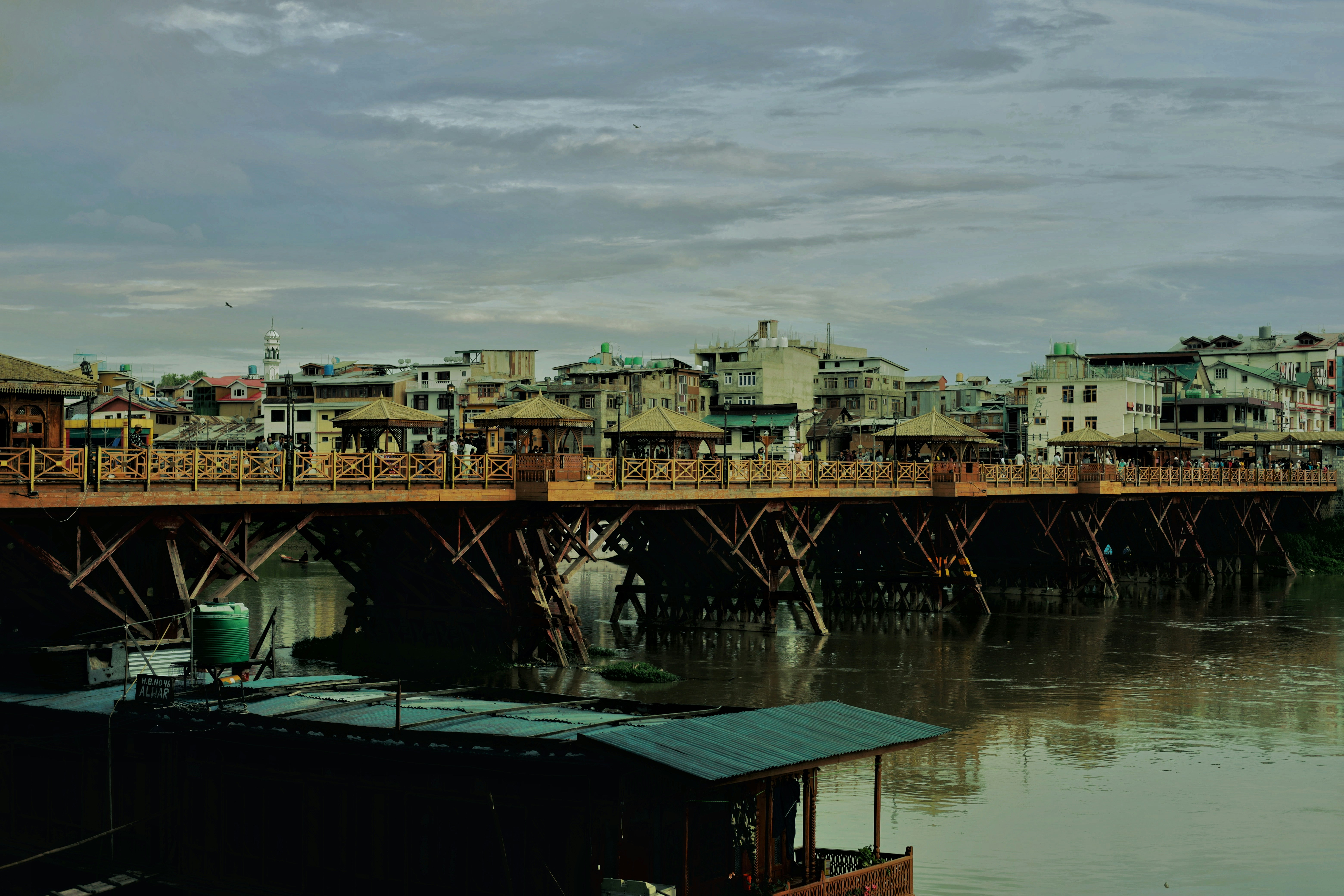 Wooden bridge spanning a reflective river, with a cluster of houses in the distance under a cloudy sky.