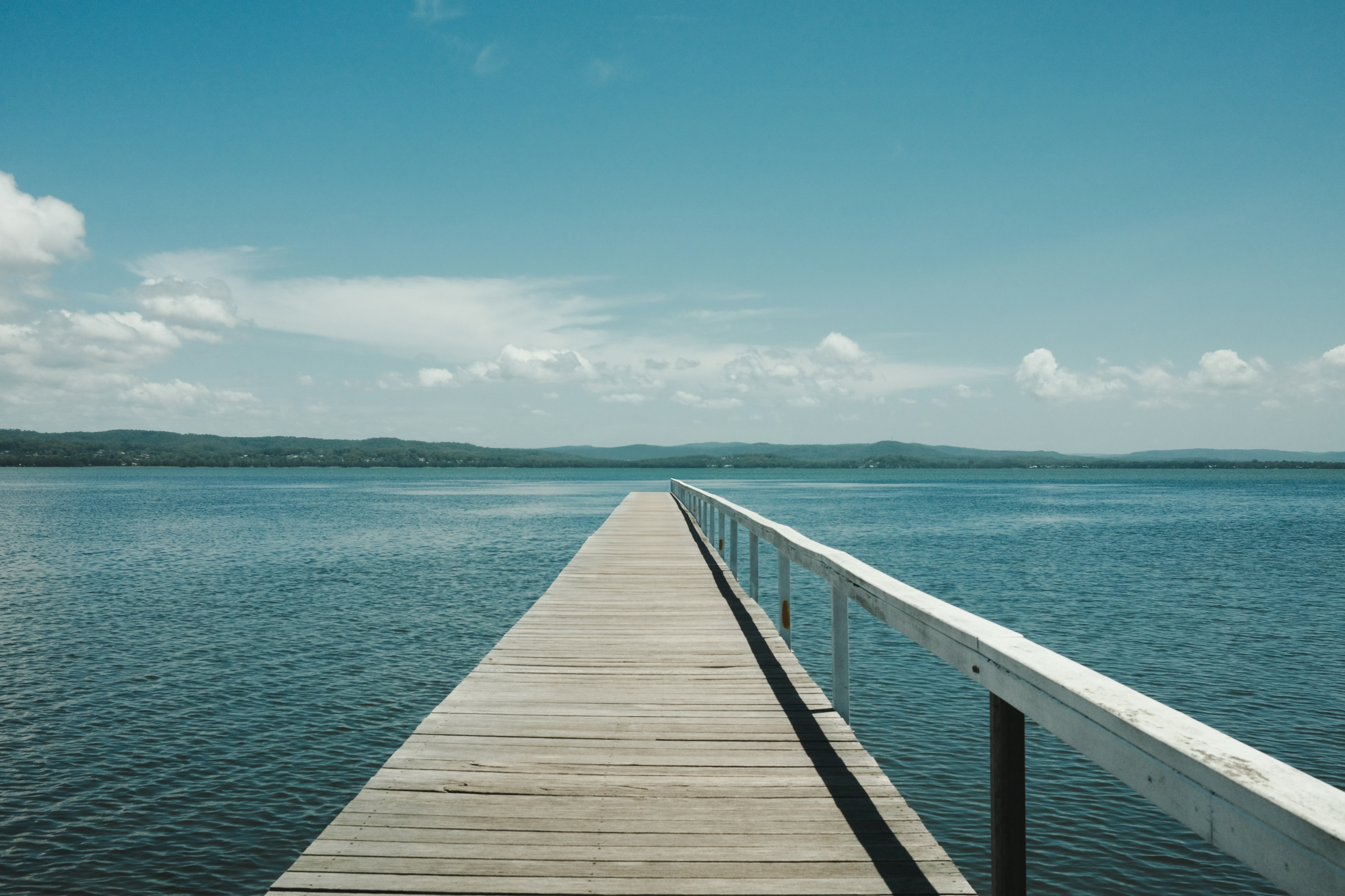 Wooden pier extends over calm blue water towards distant hills under a clear sky.