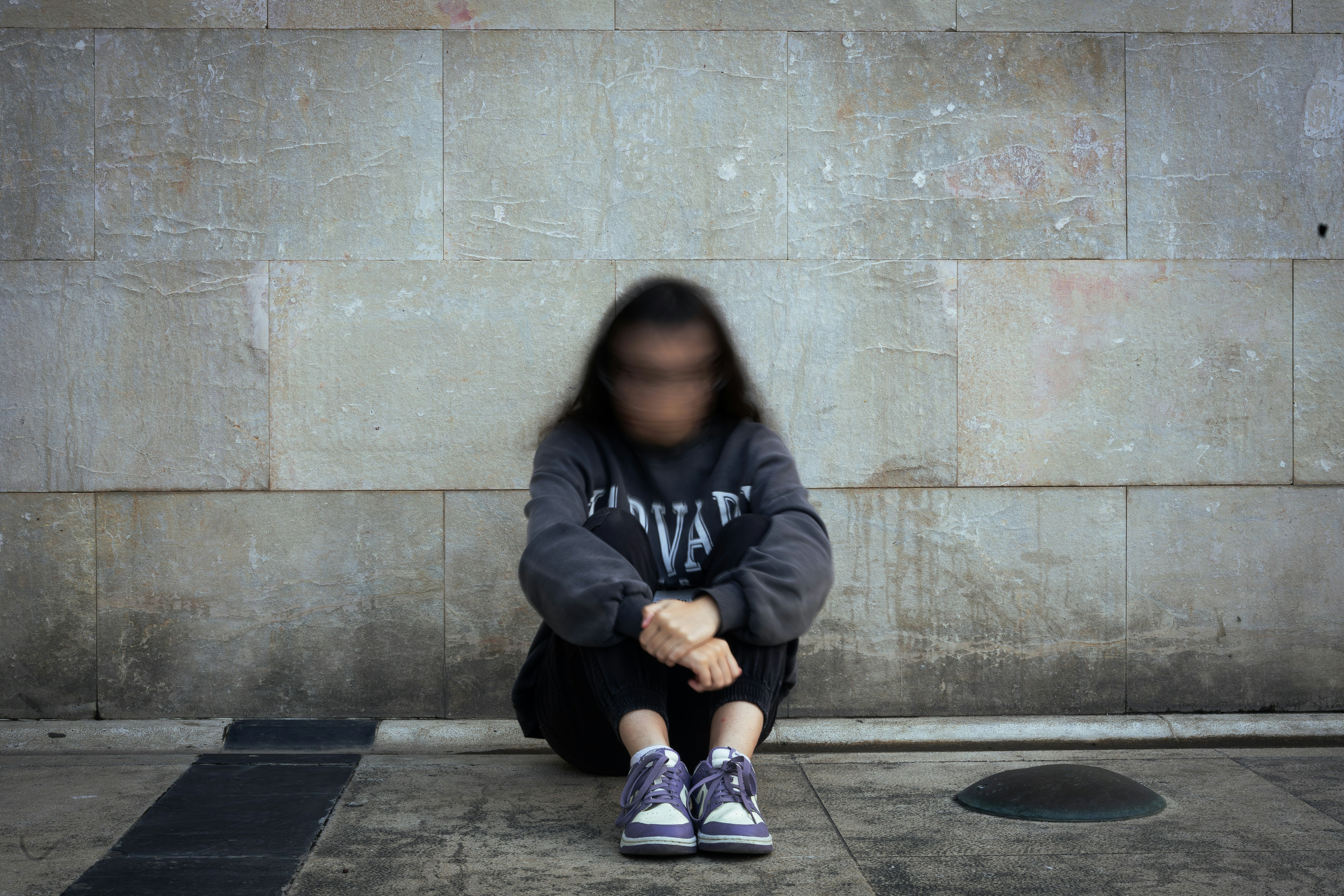 Blurred figure in a hoodie sits against a textured stone wall, creating a mood of introspection. Violet sneakers add contrast to the muted urban setting.