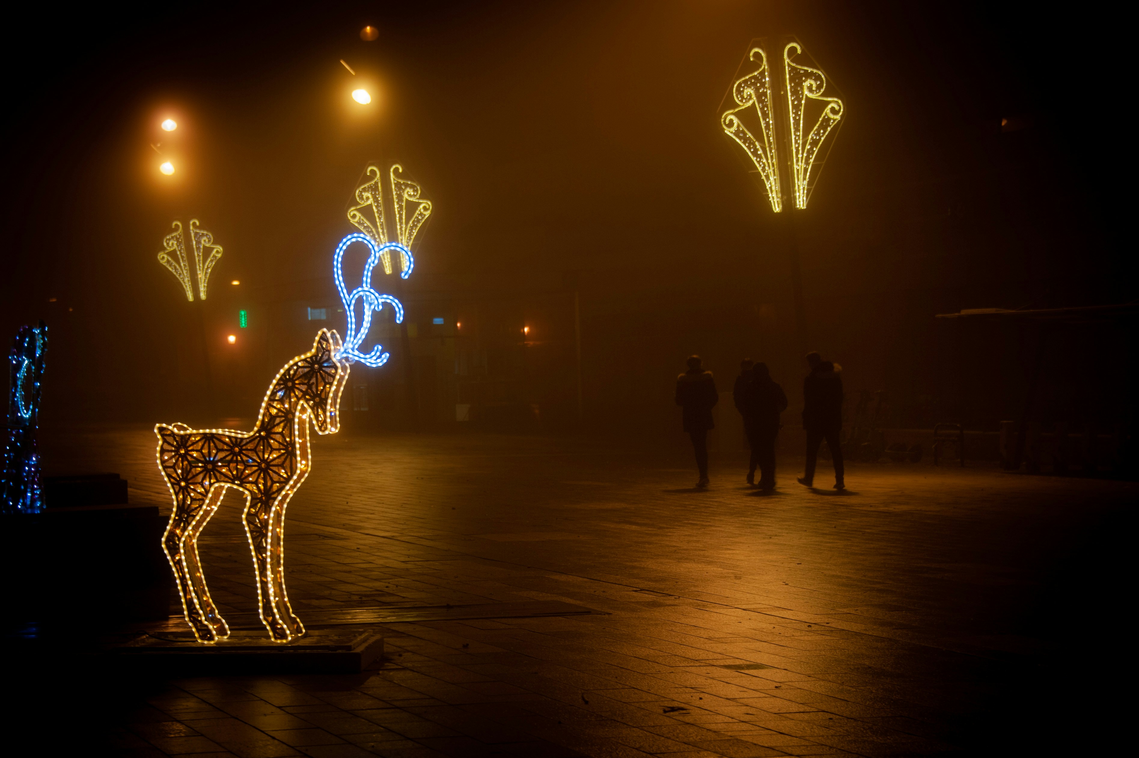Glowing reindeer sculpture with intricate lights stands in a misty urban setting, complemented by ambient streetlights and silhouetted figures.