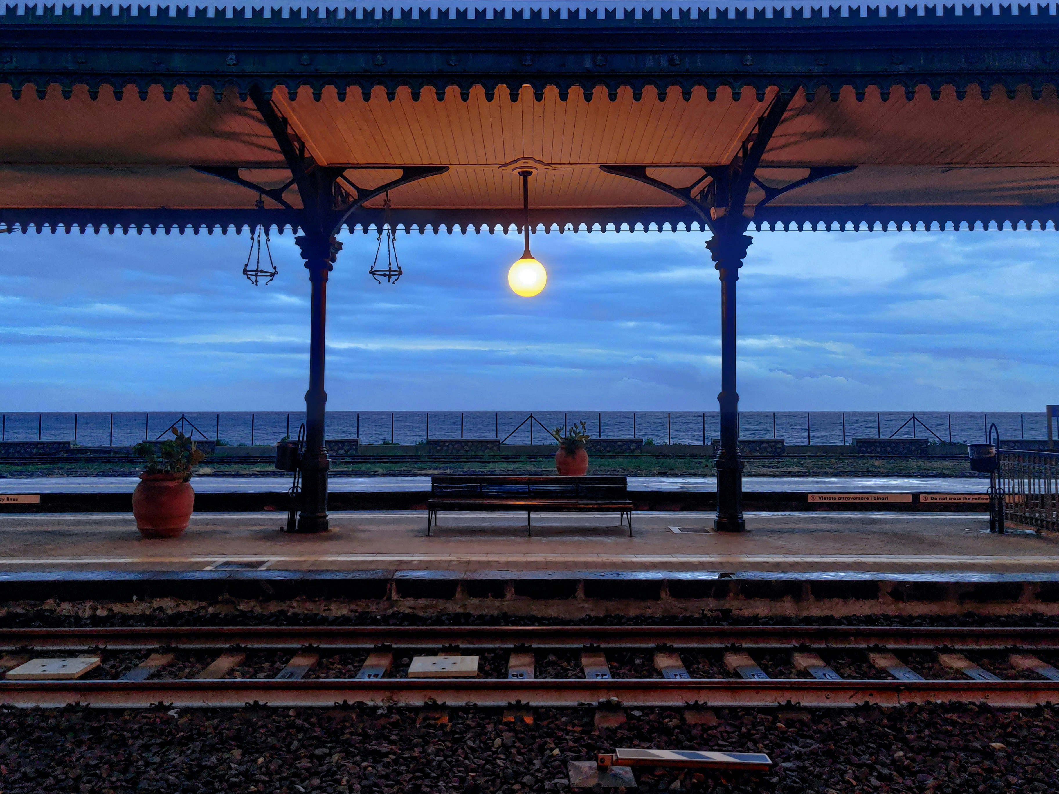 Empty train station platform beneath an ornate canopy, illuminated by a single lamp, with the ocean and twilight sky in the background.