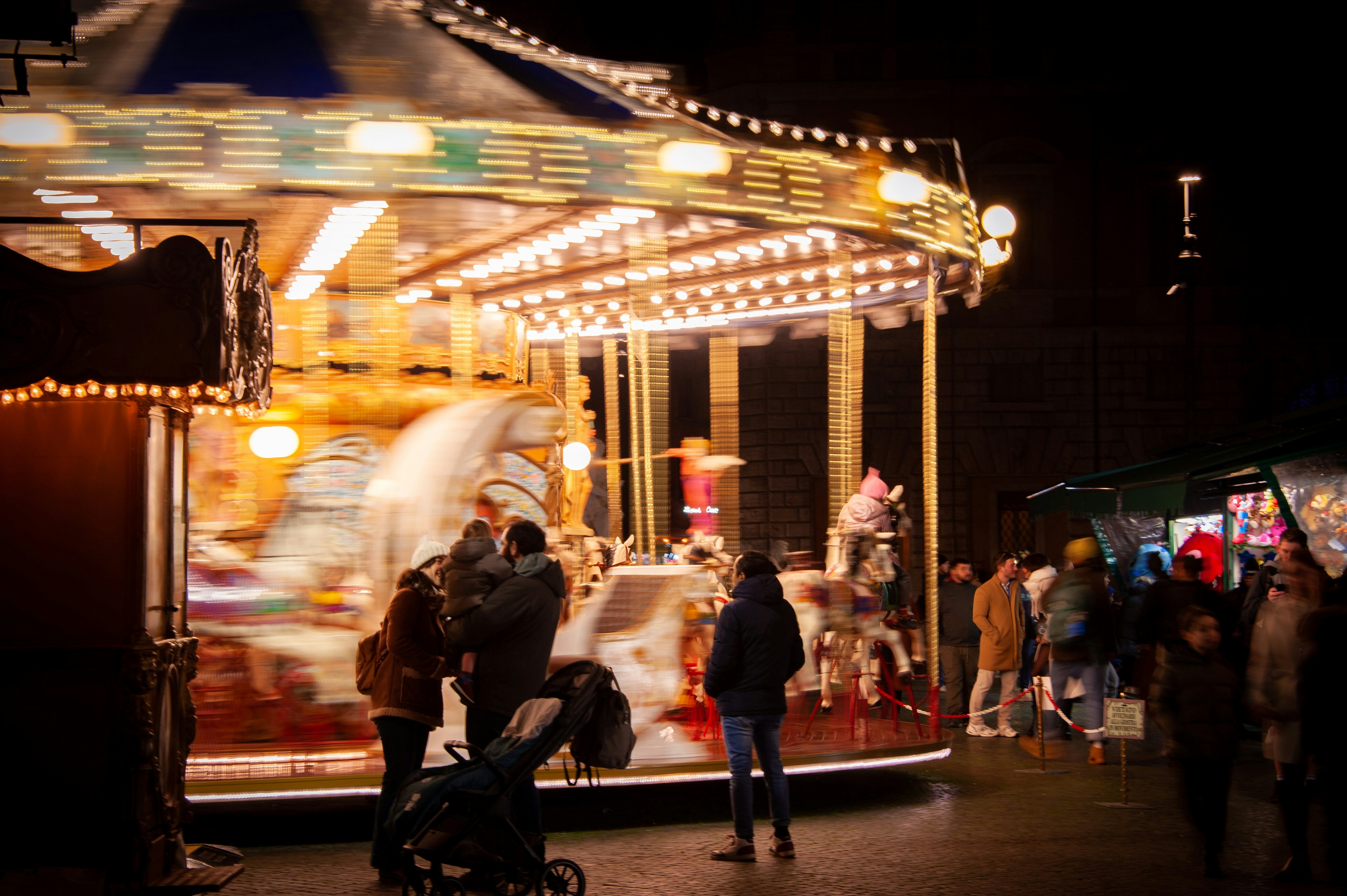 Carousel spins with vibrant lights against the night sky, surrounded by a lively crowd.