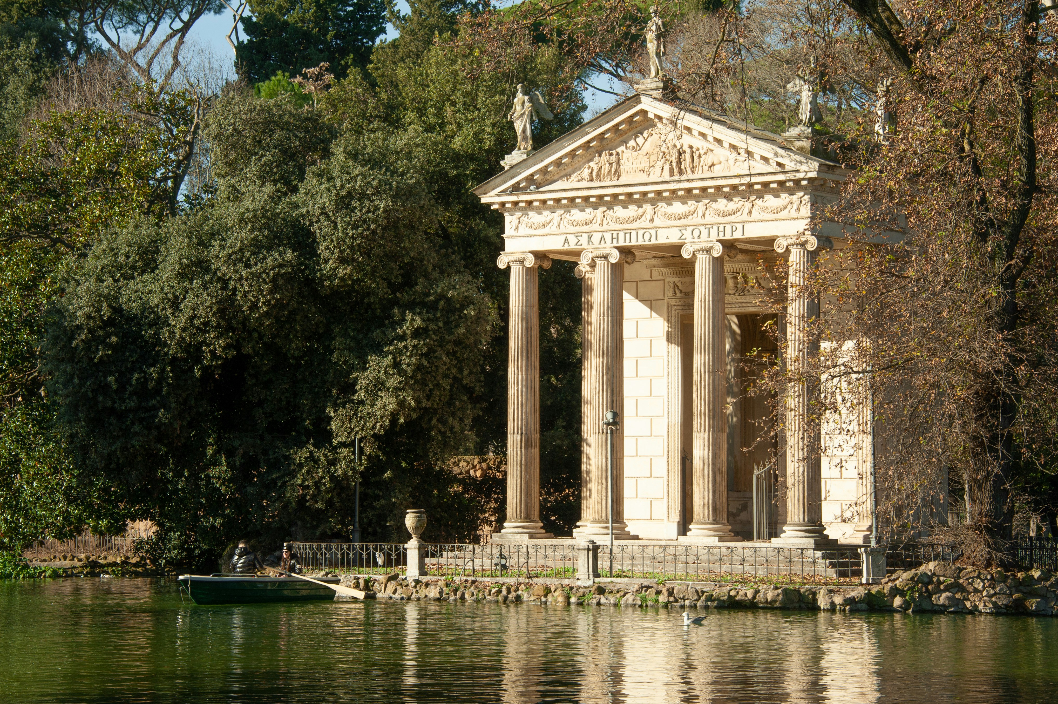 Classical temple beside a serene lake surrounded by lush greenery under warm sunlight.
