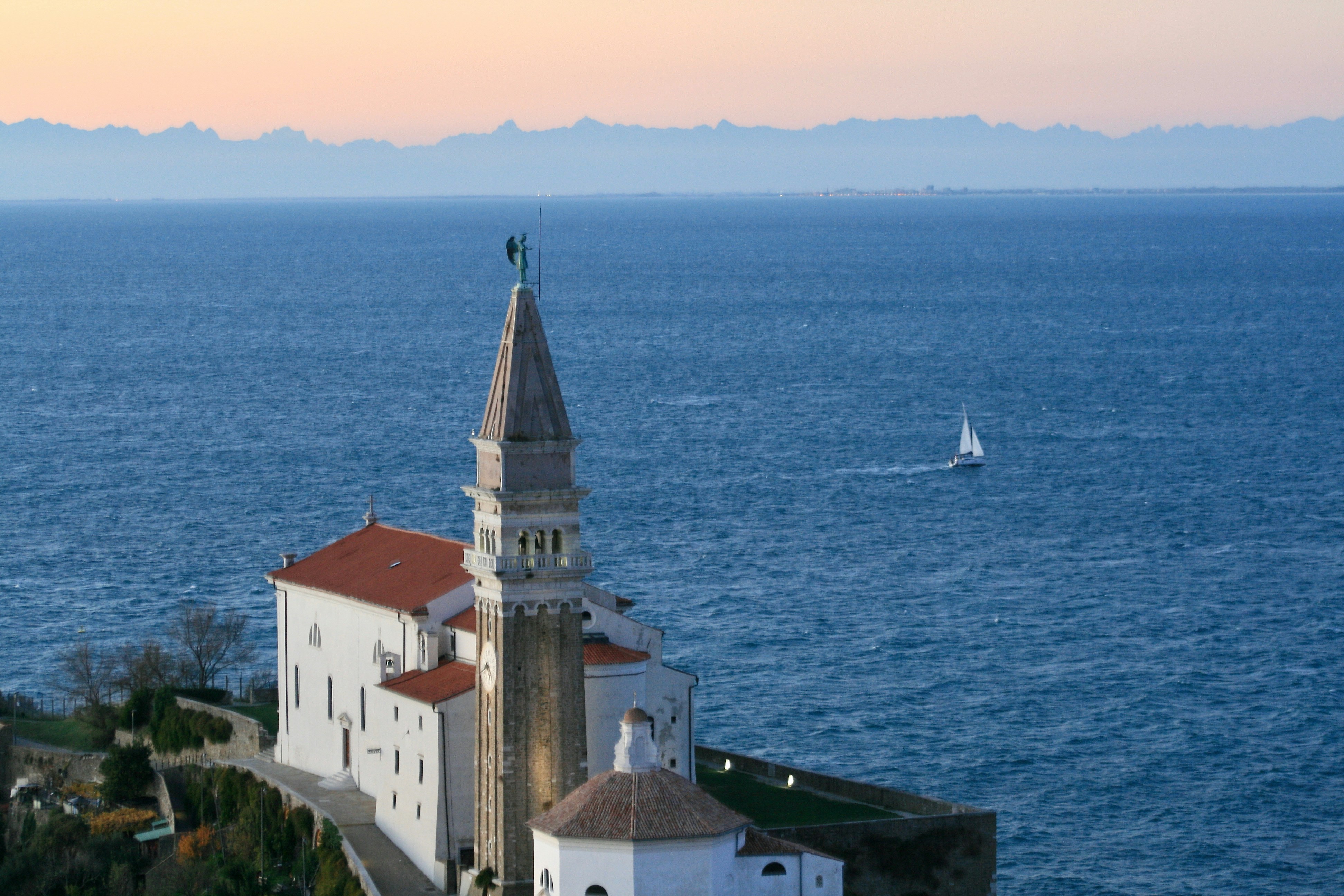 Historic coastal church overlooking a vast ocean with a distant sailboat under a pastel sunset sky.